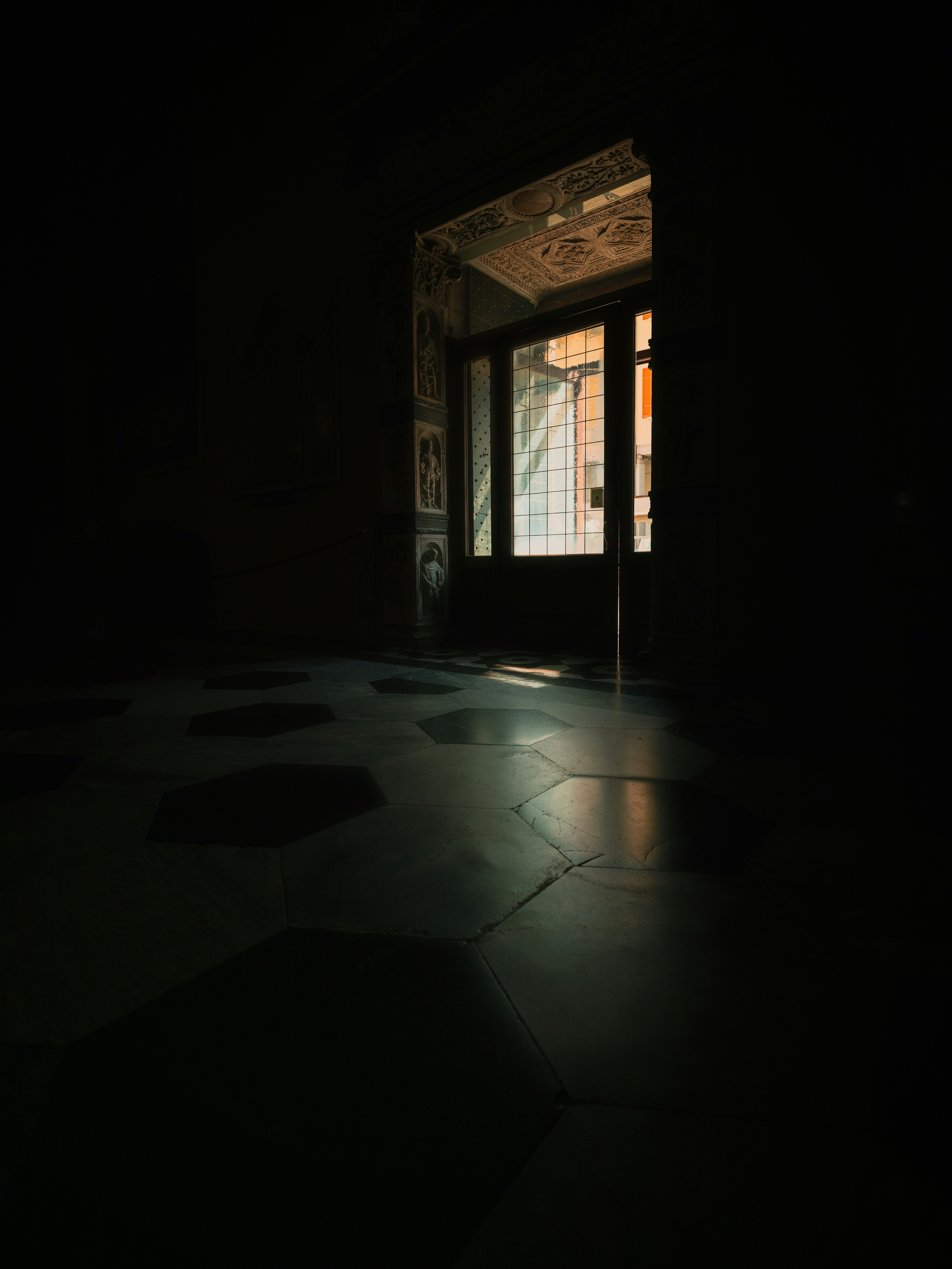 Dimly lit interior with an ornate doorway revealing a glimpse of light from outside. The patterned floor leads the eye towards the entrance.
