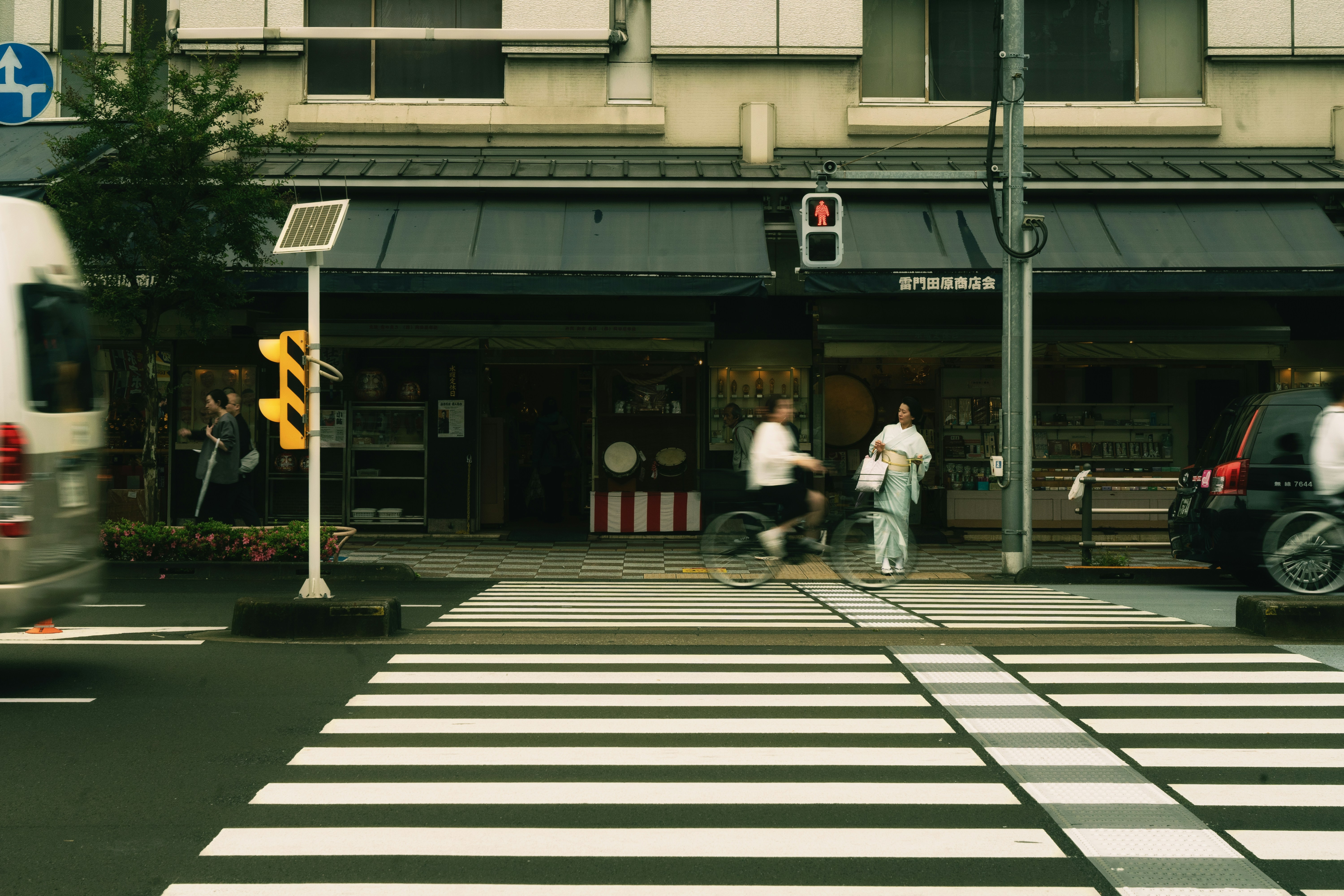 Bicyclist navigating a busy crosswalk in a vibrant urban setting, with pedestrians and vehicles in motion. The scene captures the rhythm of city life.