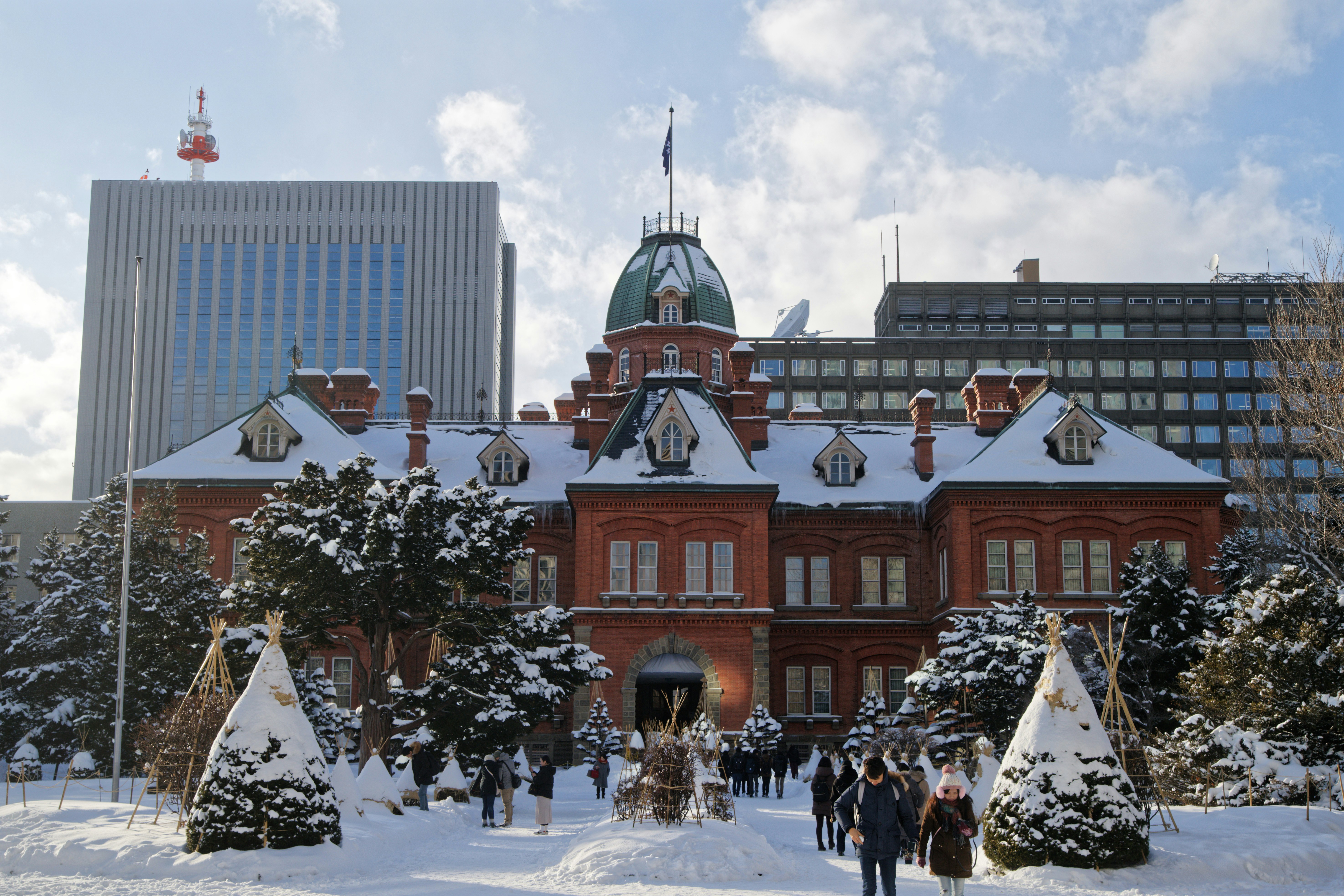 Historic red brick building surrounded by snow-covered trees and visitors, set against a backdrop of modern architecture. The scene captures the blend of history and contemporary life.