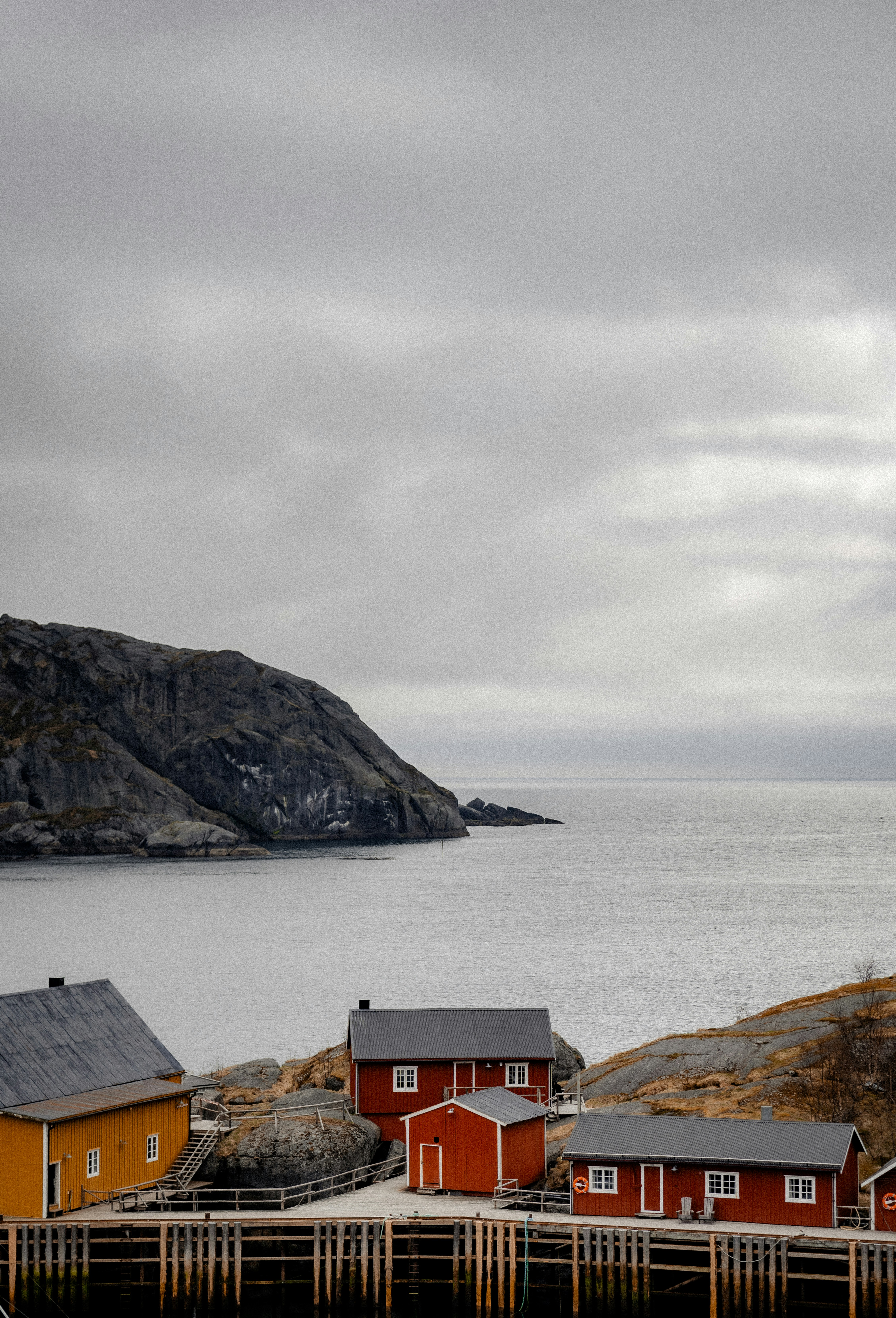Colorful wooden houses dot the shoreline beneath a cloudy sky, with rocky cliffs rising in the background. The tranquil sea reflects the muted tones of the environment.