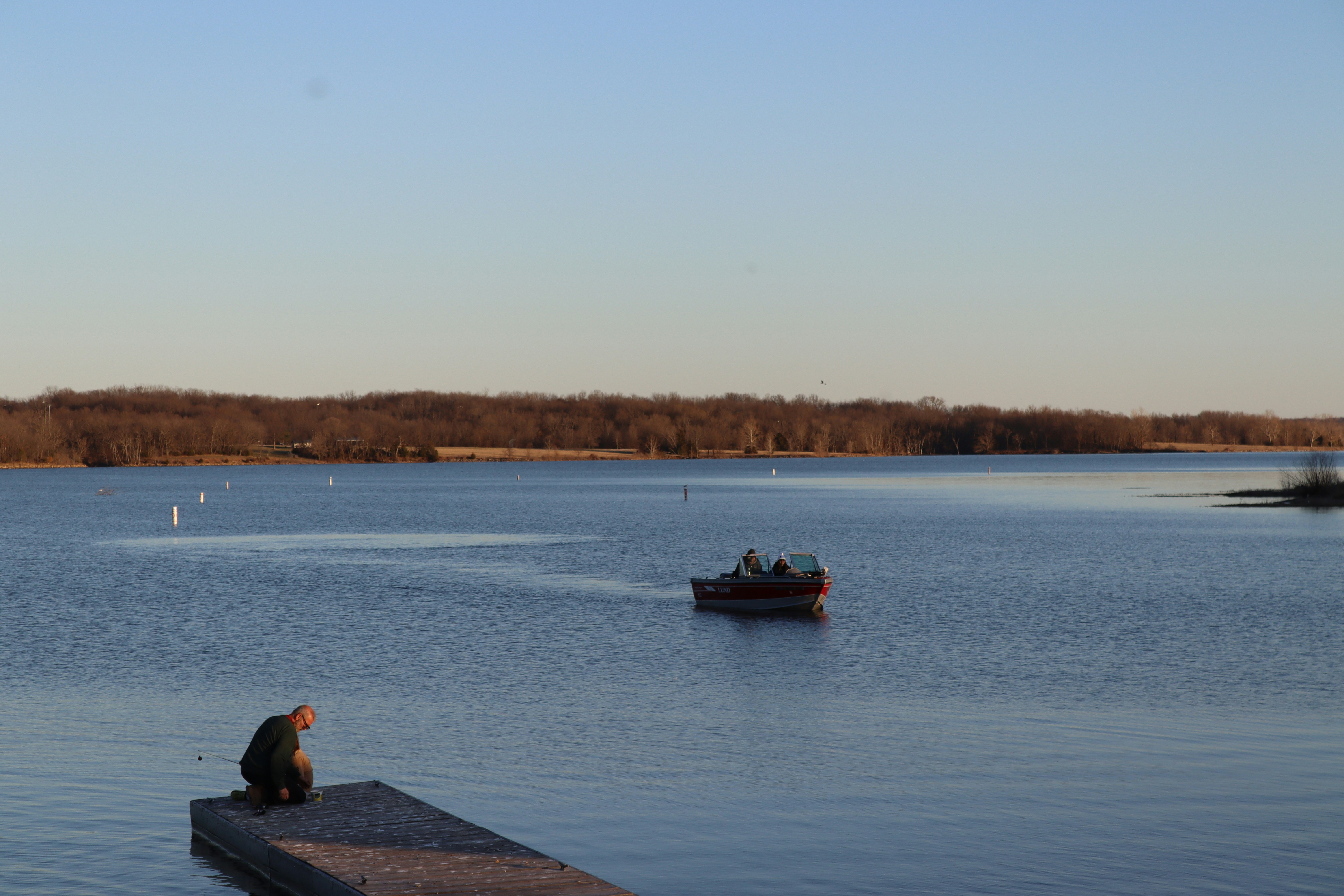 A lone figure sits on a dock, gazing at a boat navigating the calm waters of a lake, framed by a tranquil landscape.