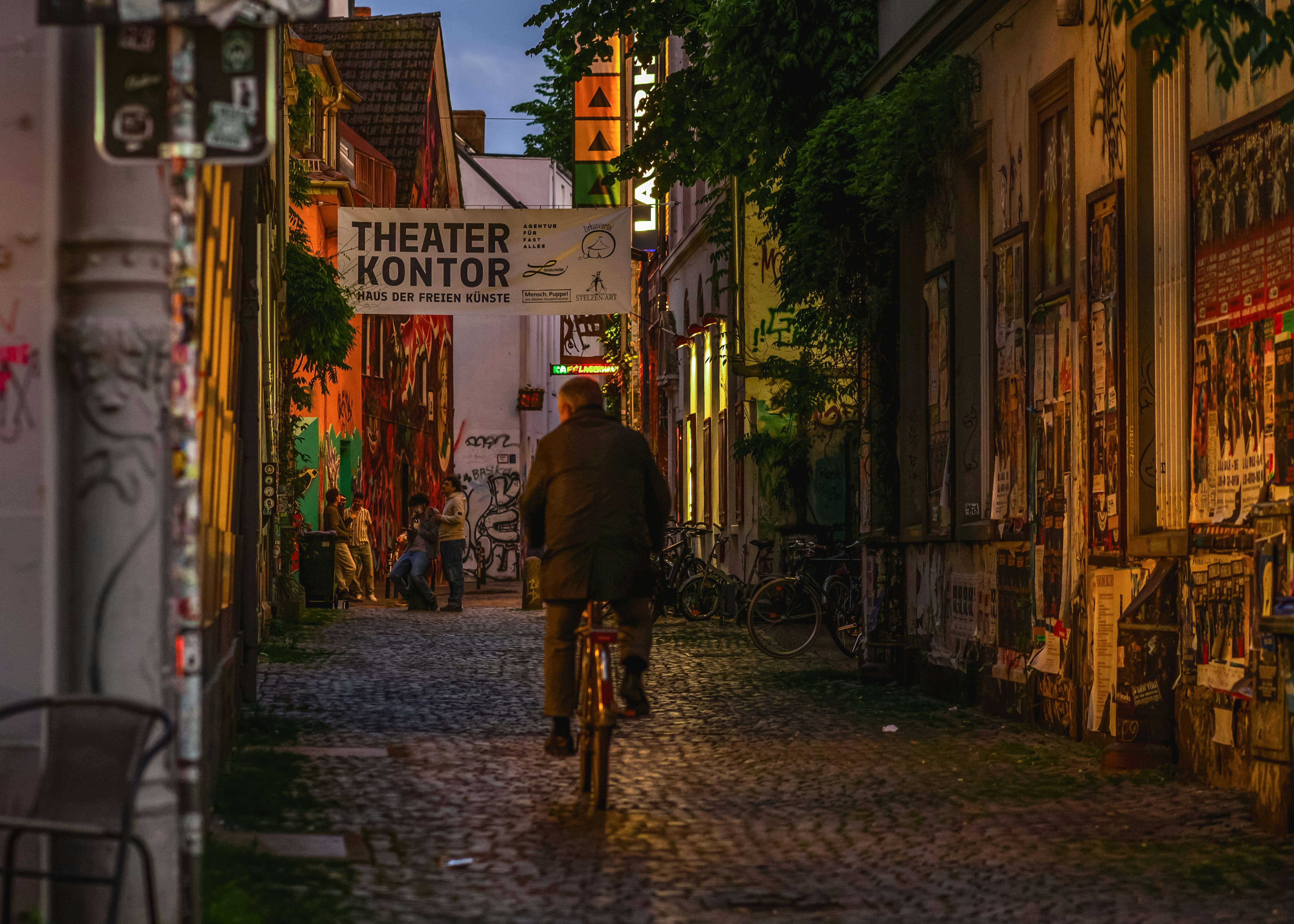 A cyclist navigates a cobblestone street adorned with vibrant murals and illuminated signs, capturing the essence of urban creativity at twilight.