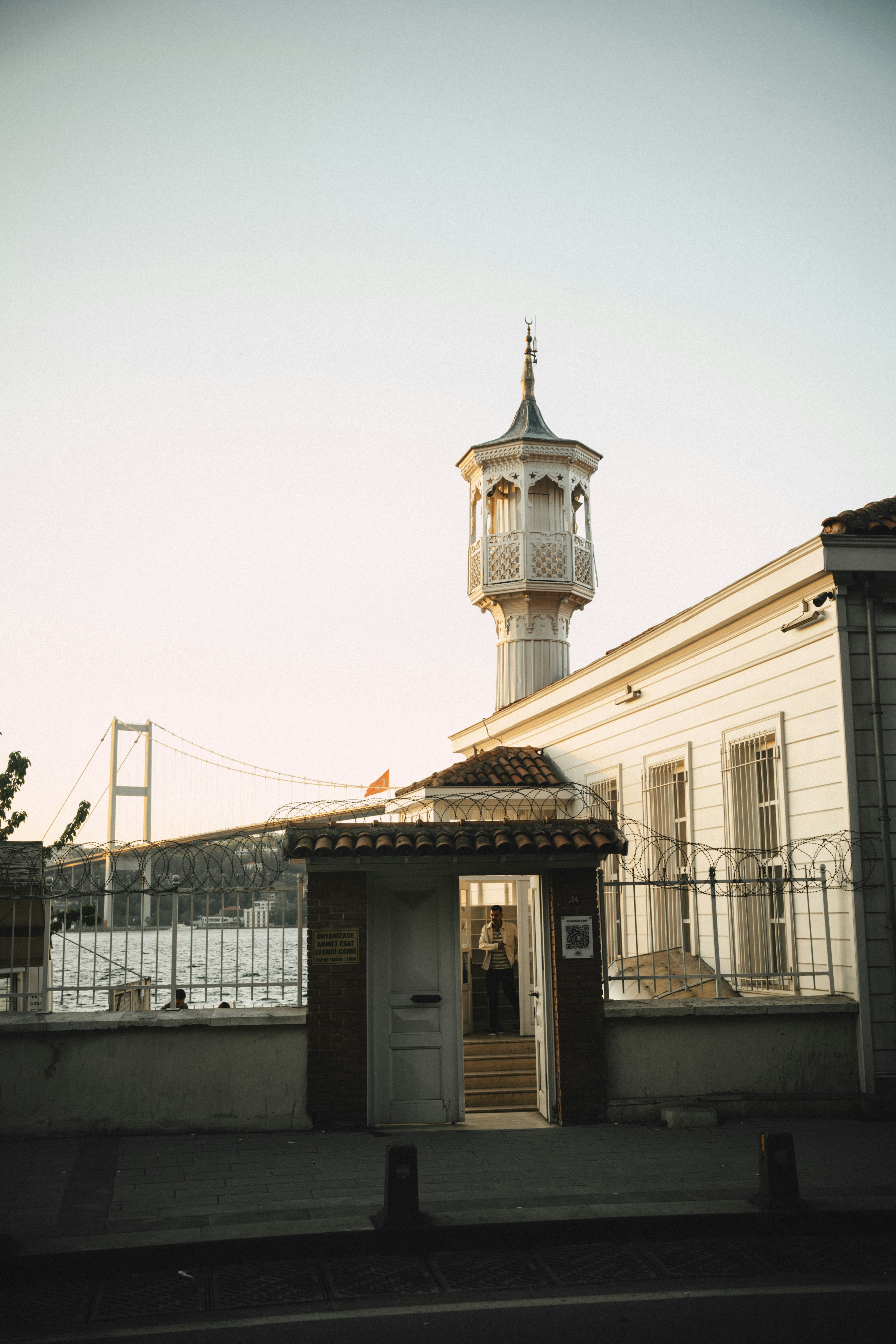 A charming waterfront building with a decorative tower, overlooking the Bosphorus Bridge at dusk. The scene captures the essence of serene urban life by the water.