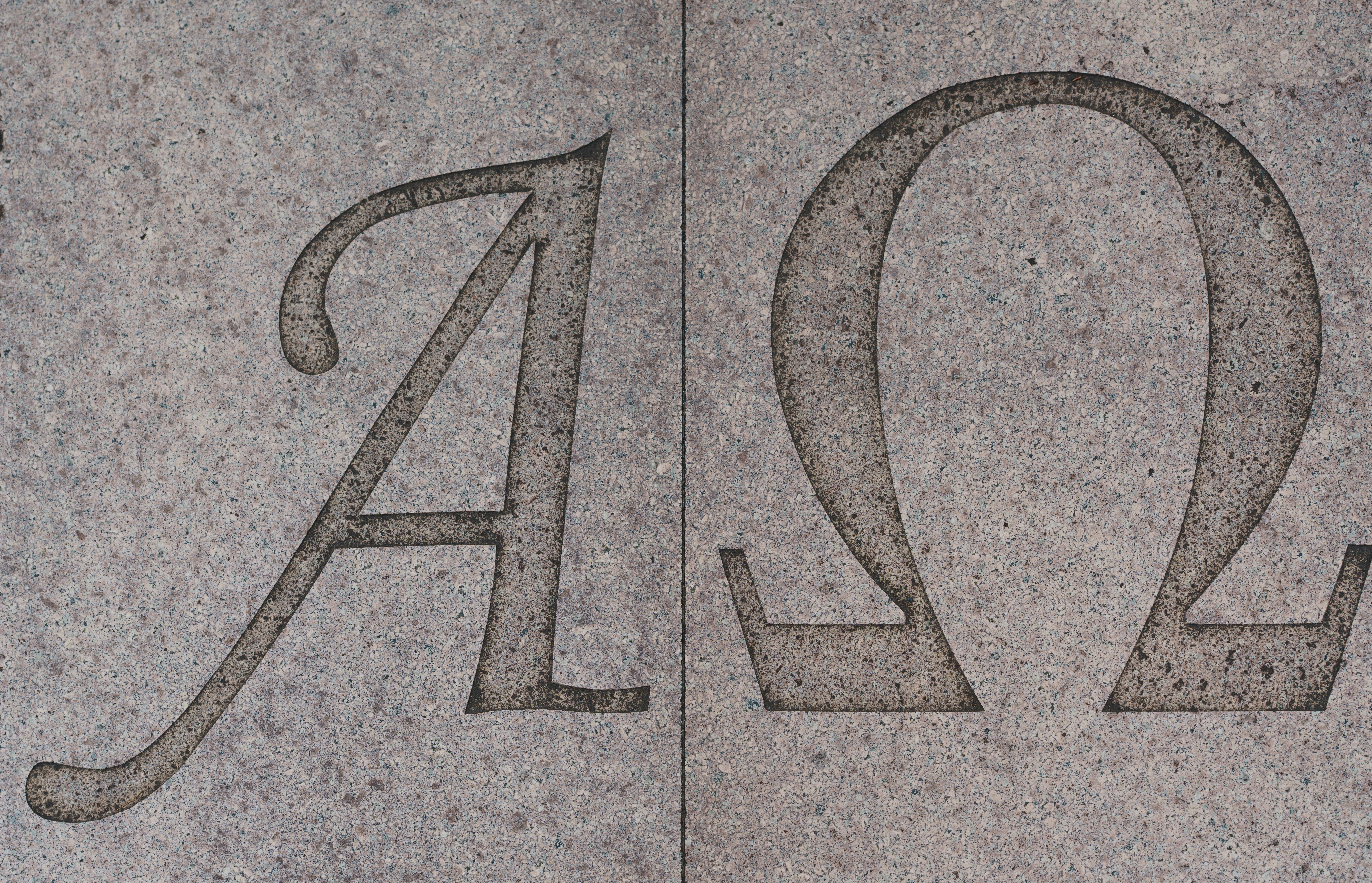 Intricate stone carvings of the letters 'A' and 'Ω' side by side, representing the concept of alpha and omega. The texture of the stone adds depth to the symbols.