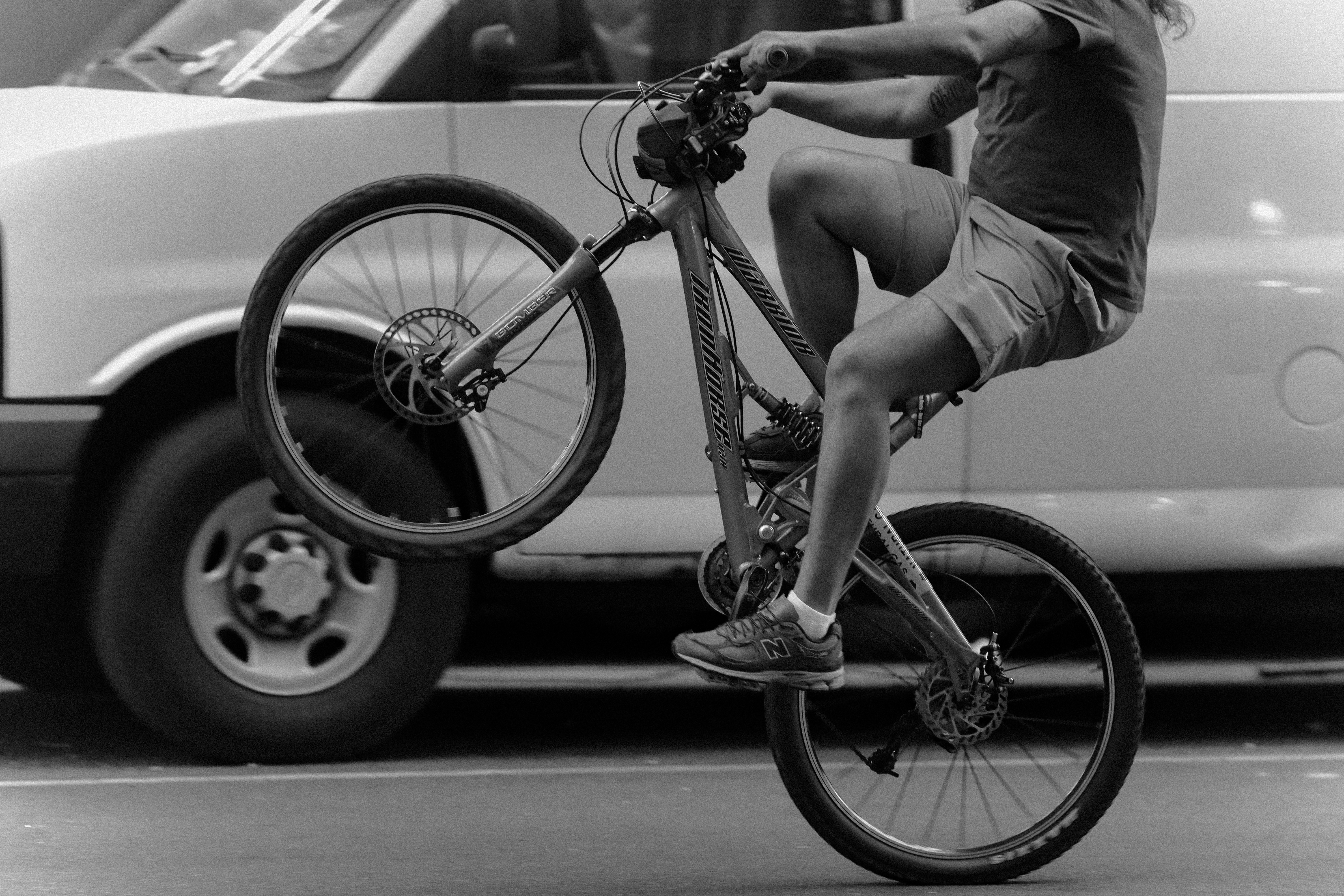 A cyclist performs a wheelie on a busy street, showcasing skill and confidence amidst the urban backdrop. The monochrome palette emphasizes the action and movement.