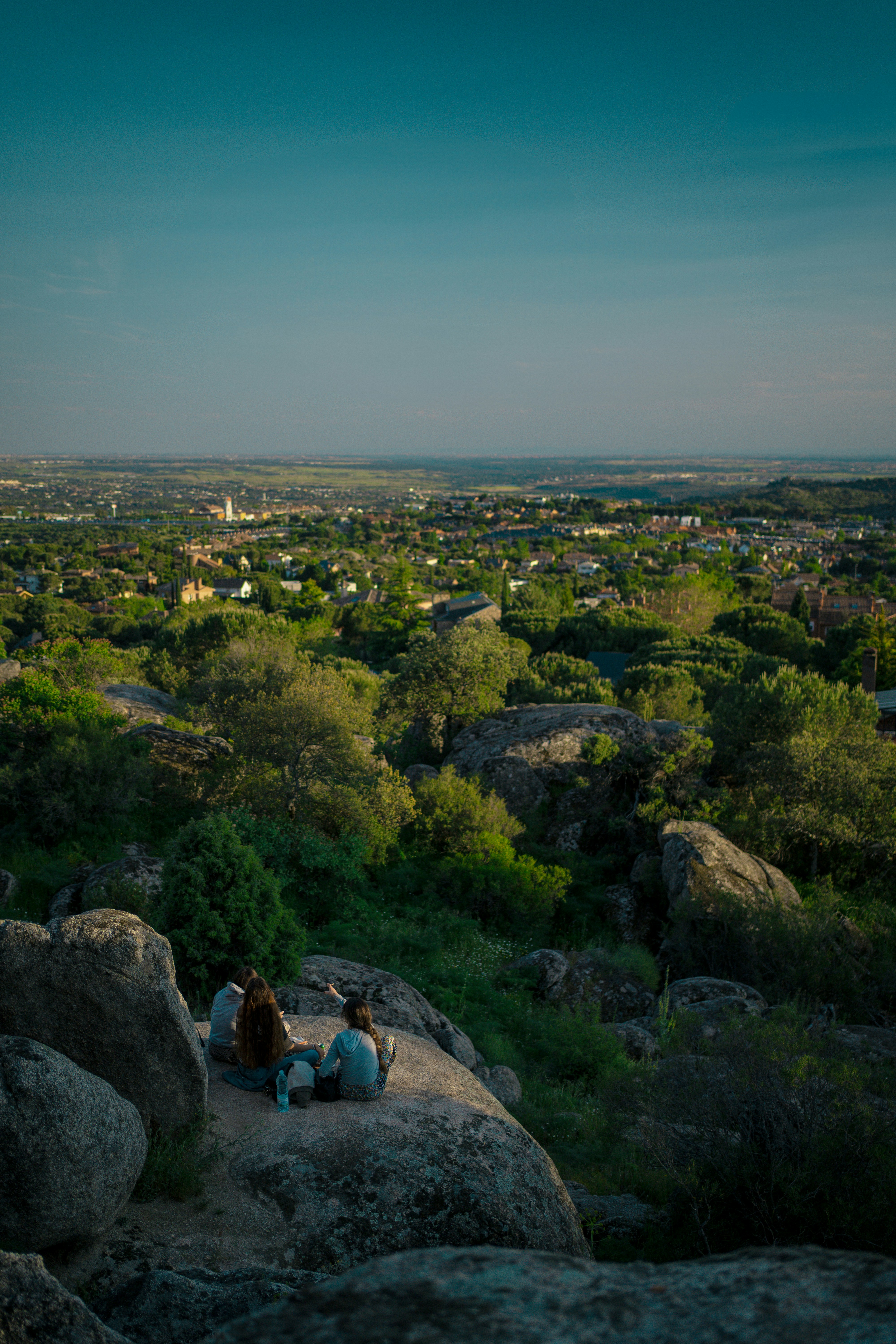 Two individuals seated on a rocky outcrop, overlooking a lush valley dotted with trees and buildings during golden hour.