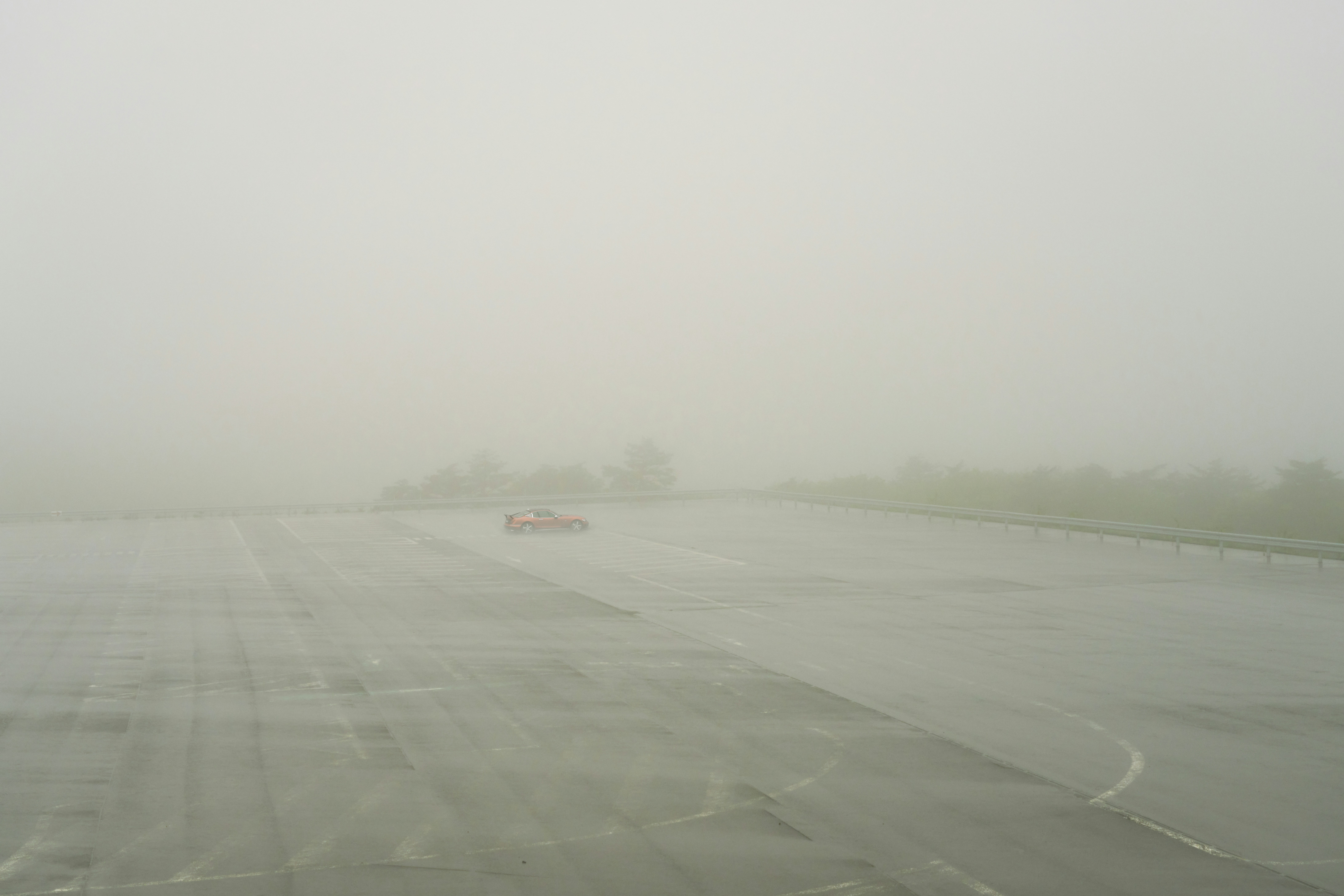 A lone red car sits on an empty, rain-soaked track shrouded in dense fog, creating an atmosphere of isolation and mystery.