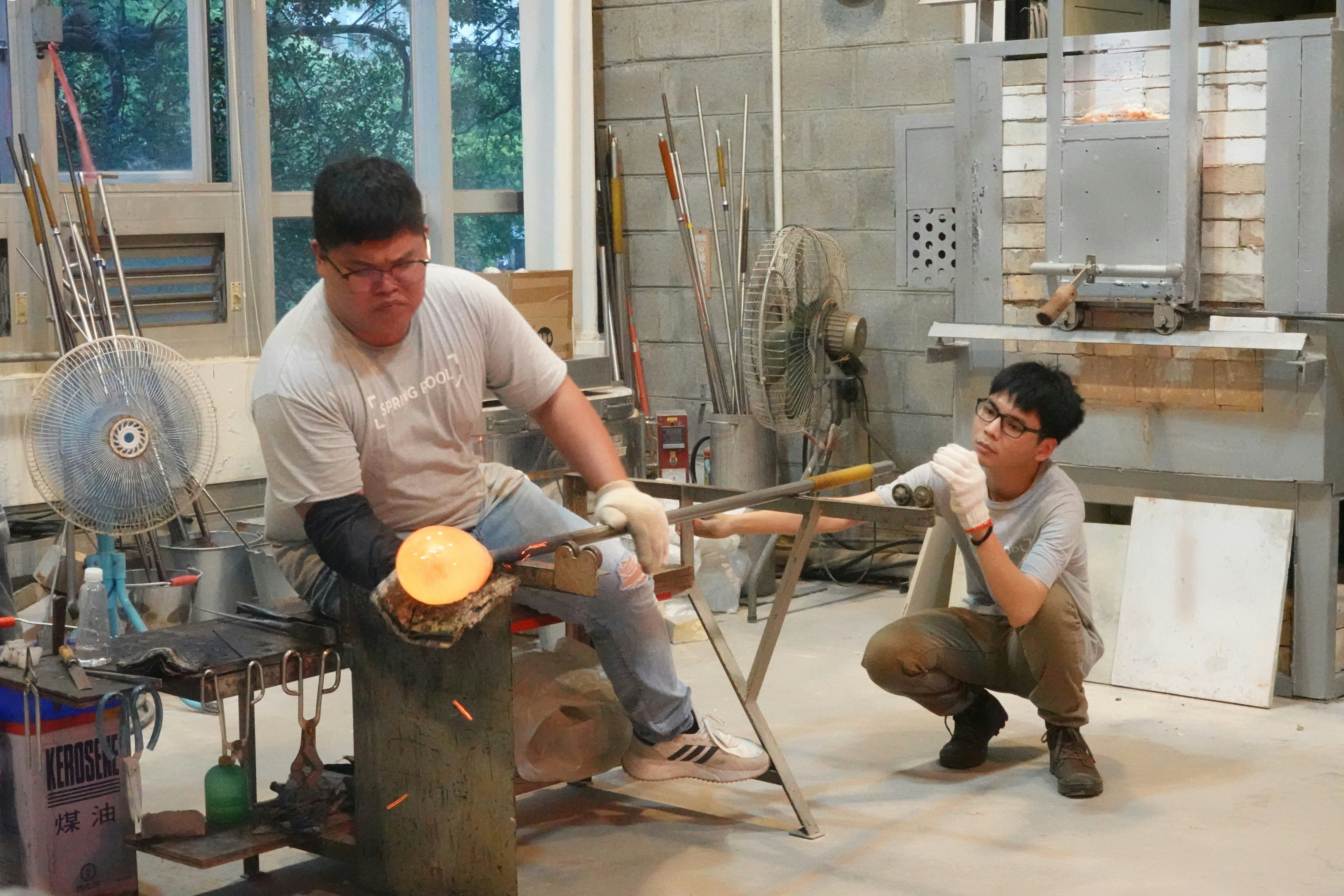 Two artisans engaged in glassblowing, with one shaping molten glass while the other assists. The workshop environment showcases tools and equipment essential for the craft.