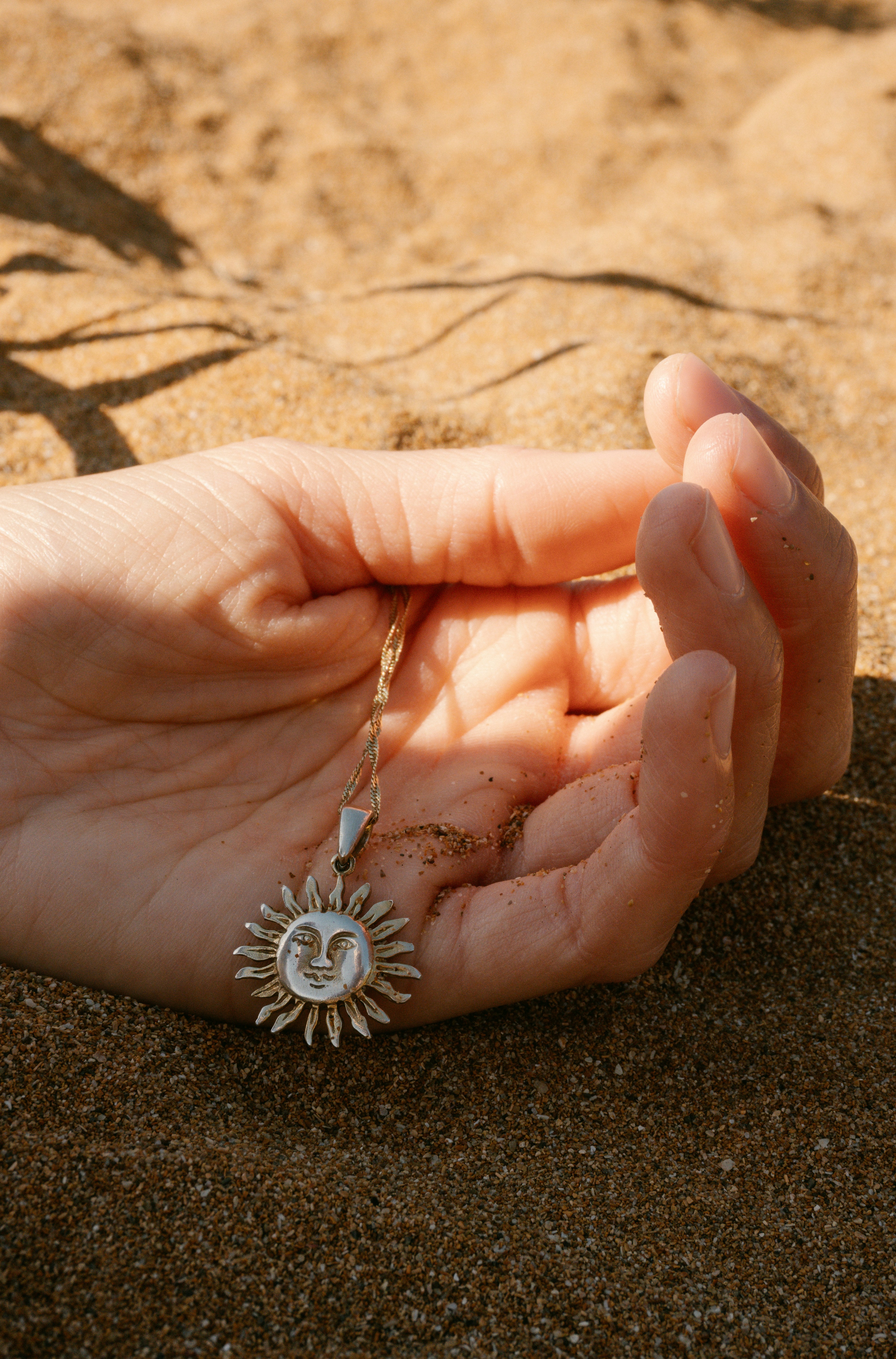 Delicate silver pendant shaped like the sun rests in an outstretched hand, surrounded by soft sand. The warm glow of sunlight enhances its intricate design.