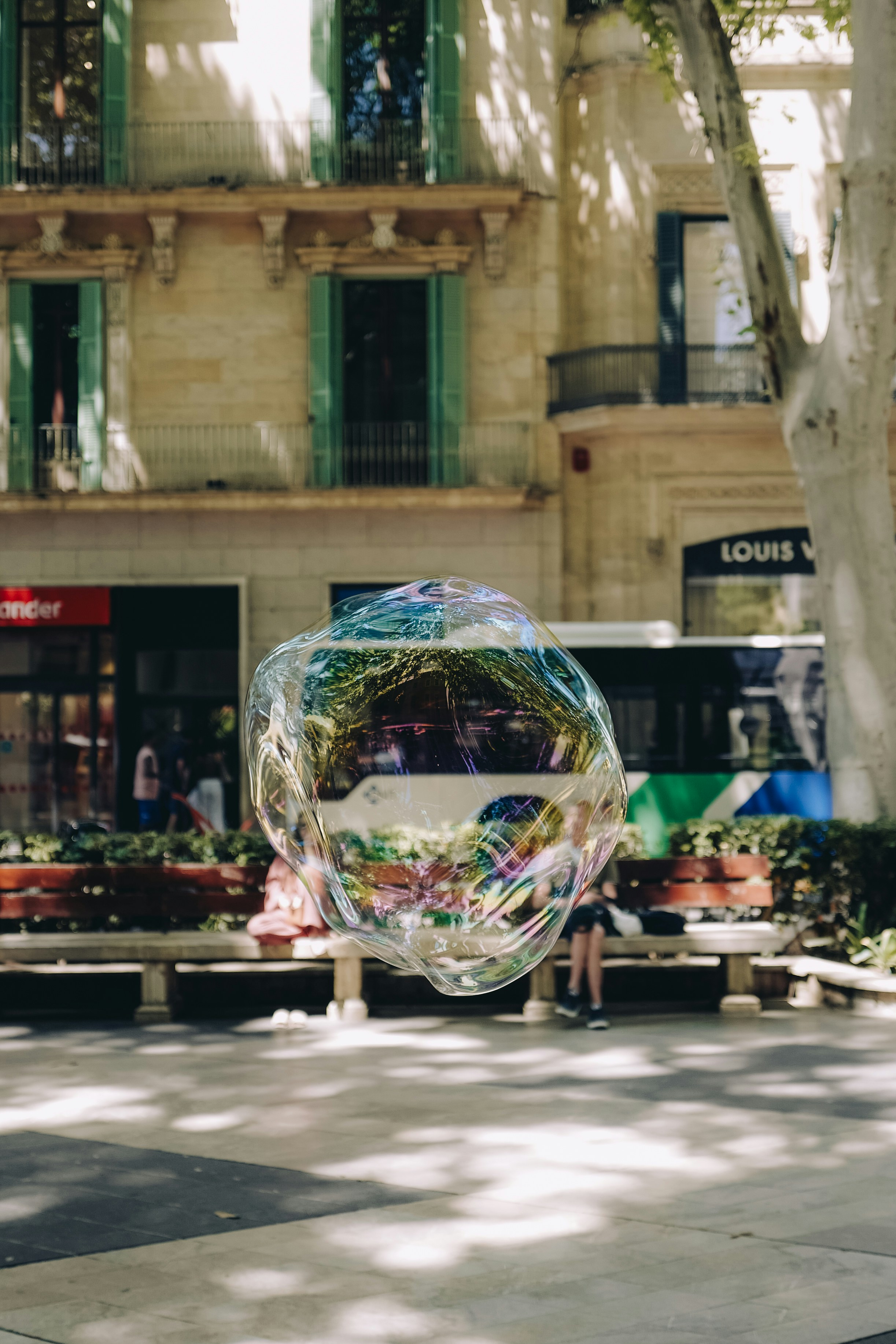 A large soap bubble floats gracefully in a sunlit plaza, capturing reflections of the surrounding buildings and trees. The scene conveys a moment of whimsy and tranquility.