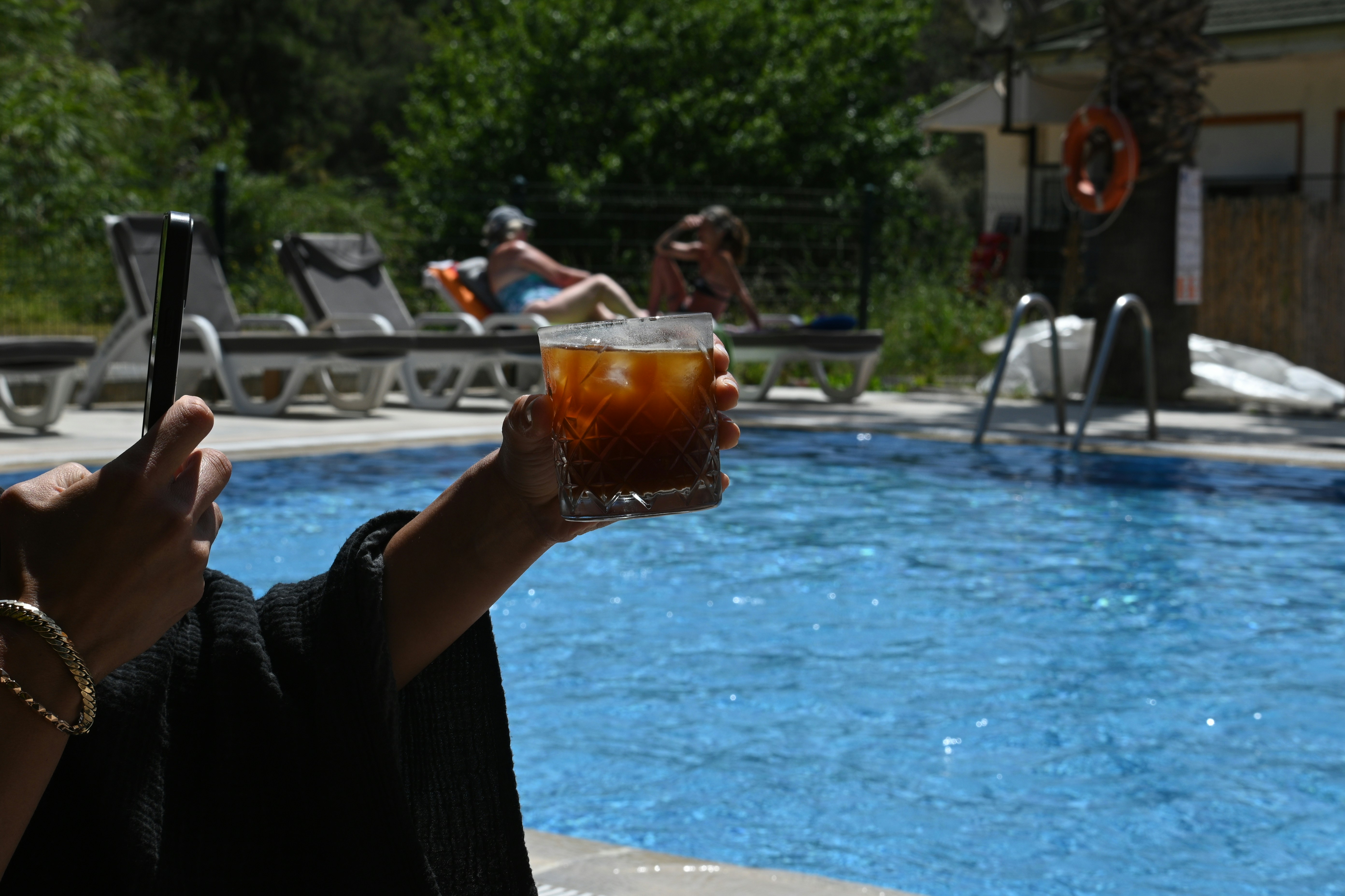 A hand holding a cocktail glass near a shimmering pool, with sunbathers relaxing in the background. The scene exudes a leisurely summer atmosphere.