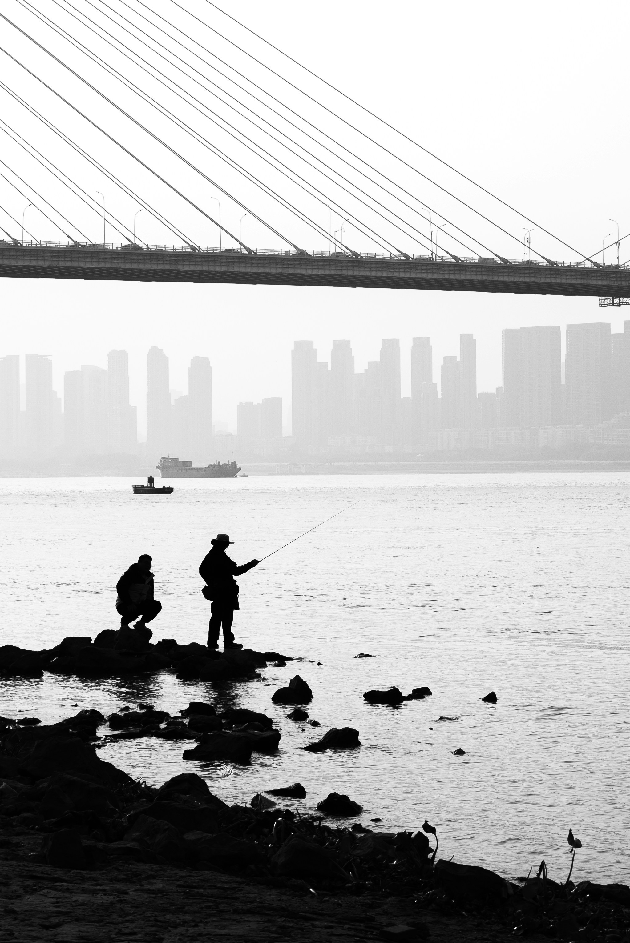 On the Rocks: Two Anglers at Dusk Under a Cable-Streaked Bridge