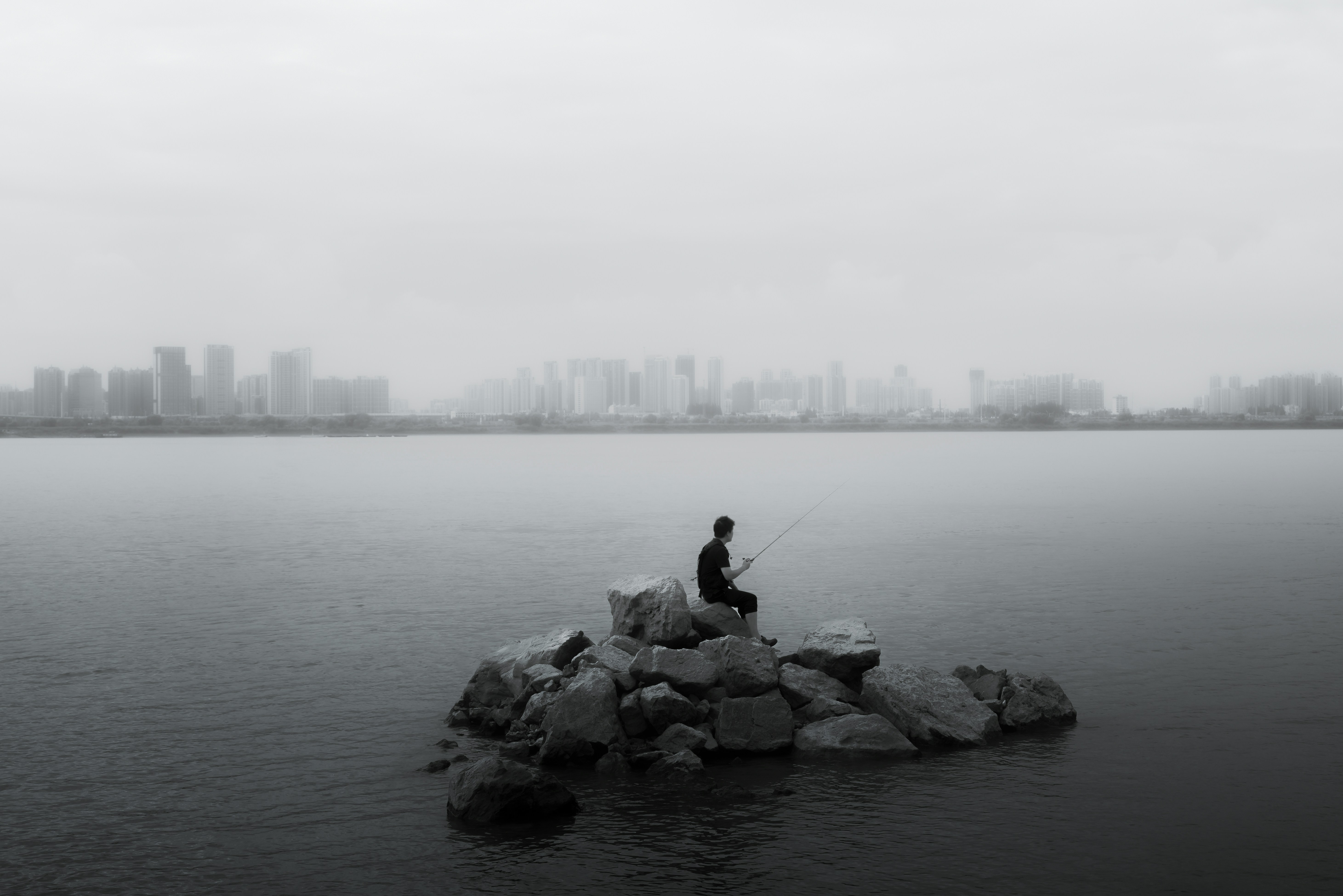 A lone fisherman sits on a rocky outcrop, casting his line into a calm body of water, with a misty city skyline in the background.