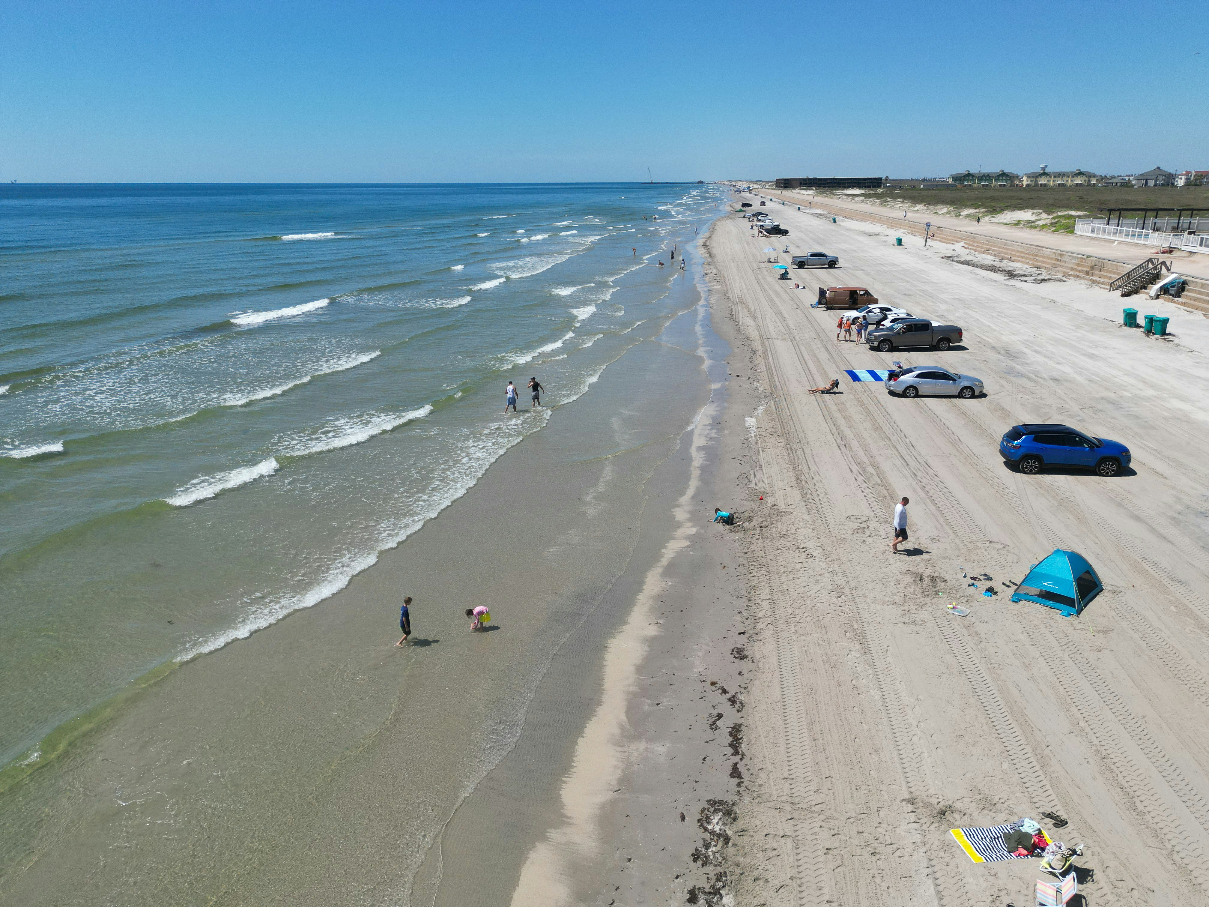 Families enjoying a sunny day on the beach, with gentle waves lapping at the shore and parked cars lining the sandy beach.
