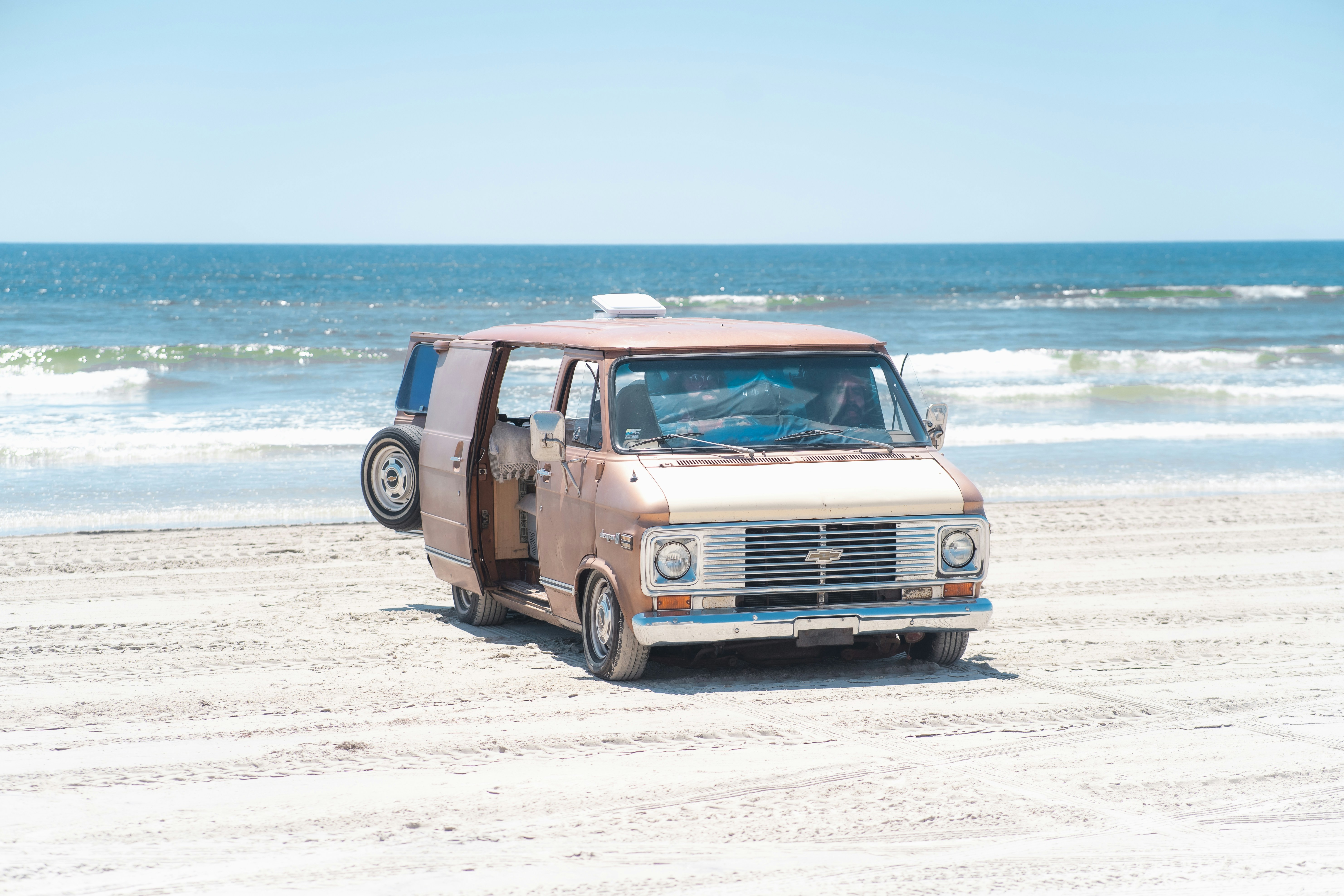 A vintage brown van, playfully nicknamed "Lil' Brown Jug," is parked on the sandy beach in Corpus Christi, its side door open. Beach chairs and a cooler sit beside it, suggesting a relaxed day by the Gulf of Mexico where people can be seen enjoying the water in the background under a bright, sunny sky.