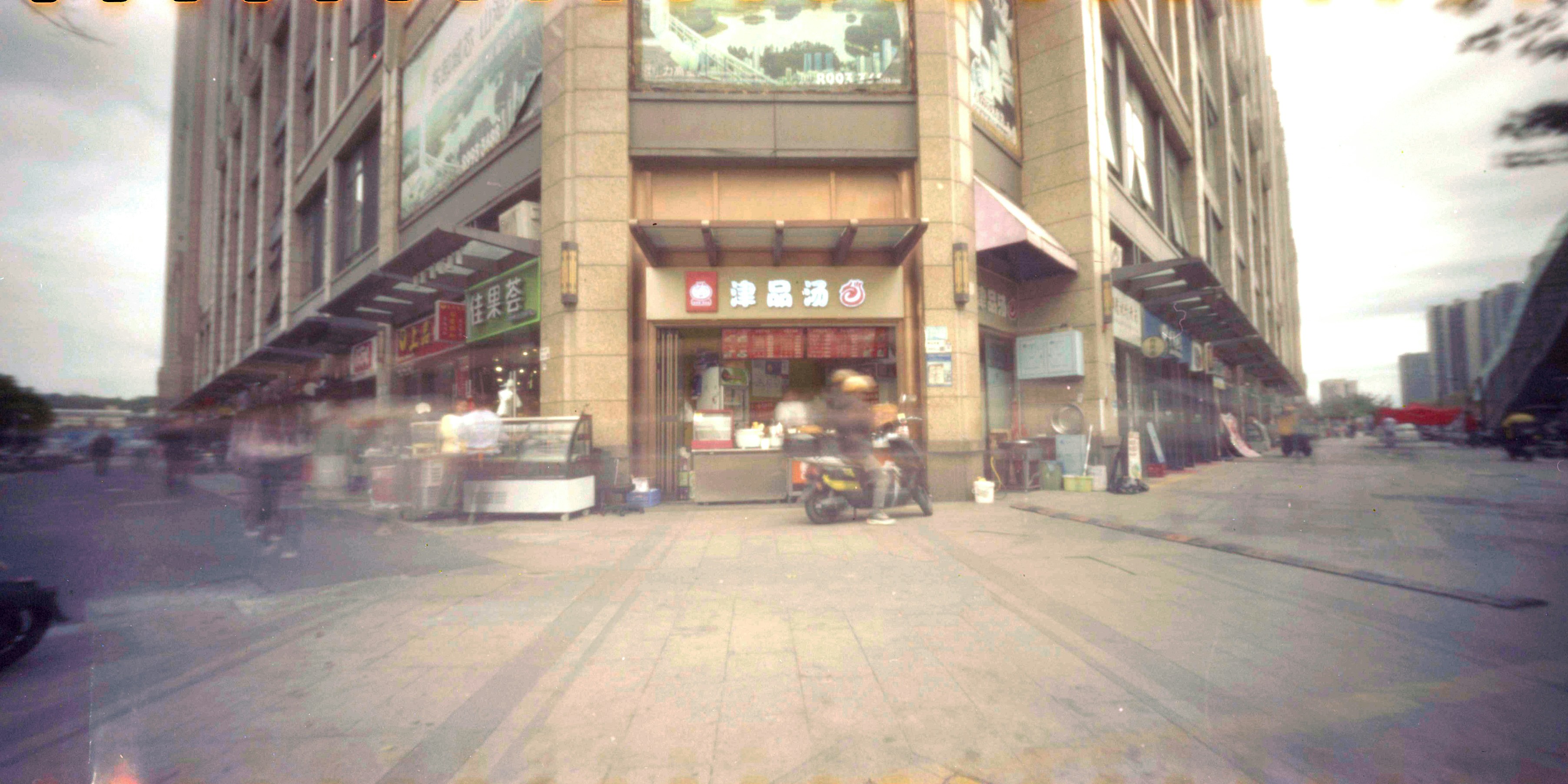 A fleeting figure pauses at a street-corner shop, makes a quick purchase, and vanishes into the urban flow. The wide-format composition crams two intersecting streets onto one frame—visually arresting.