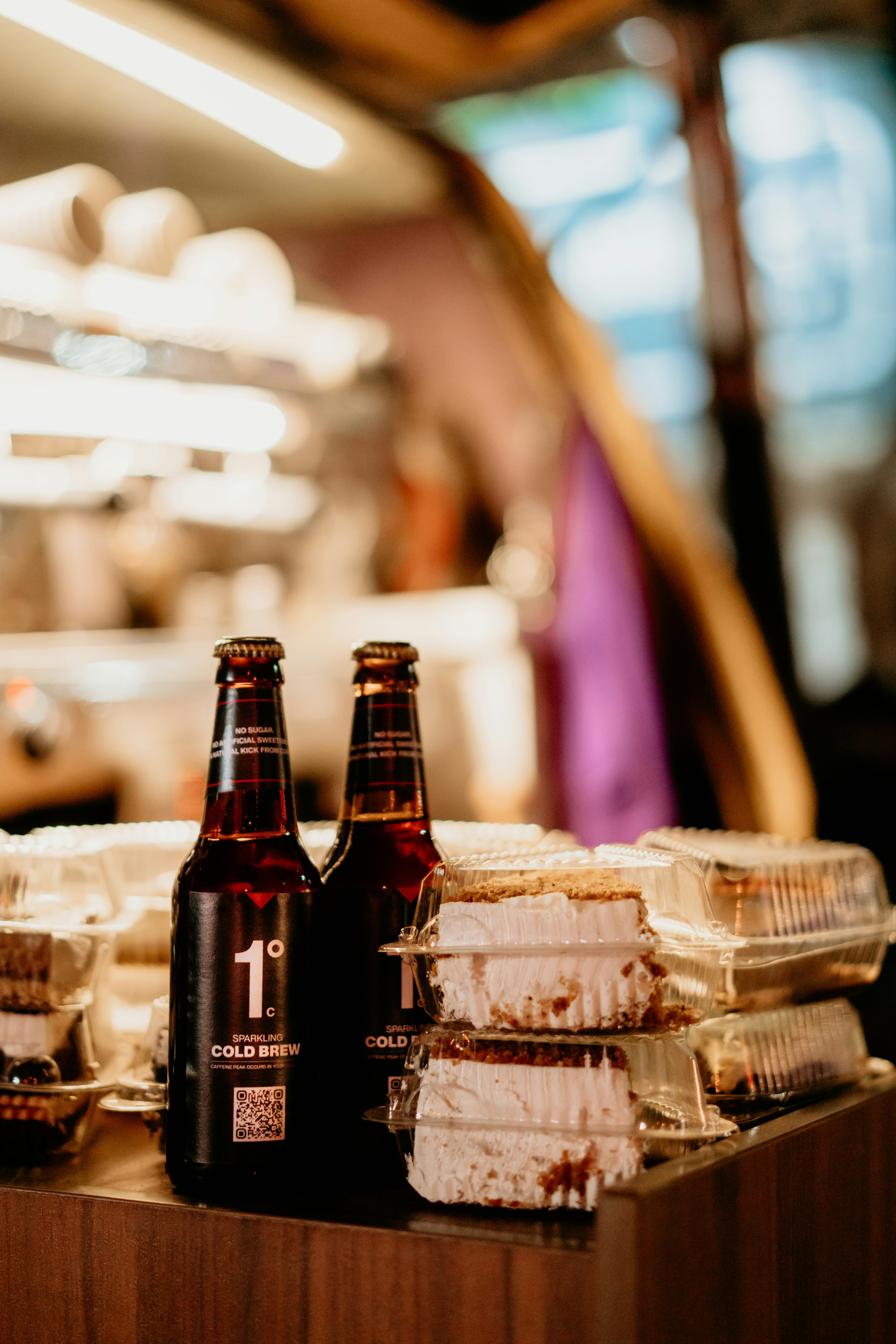 Two bottles of cold brew coffee sit prominently in front of neatly stacked dessert containers in a cozy café setting.