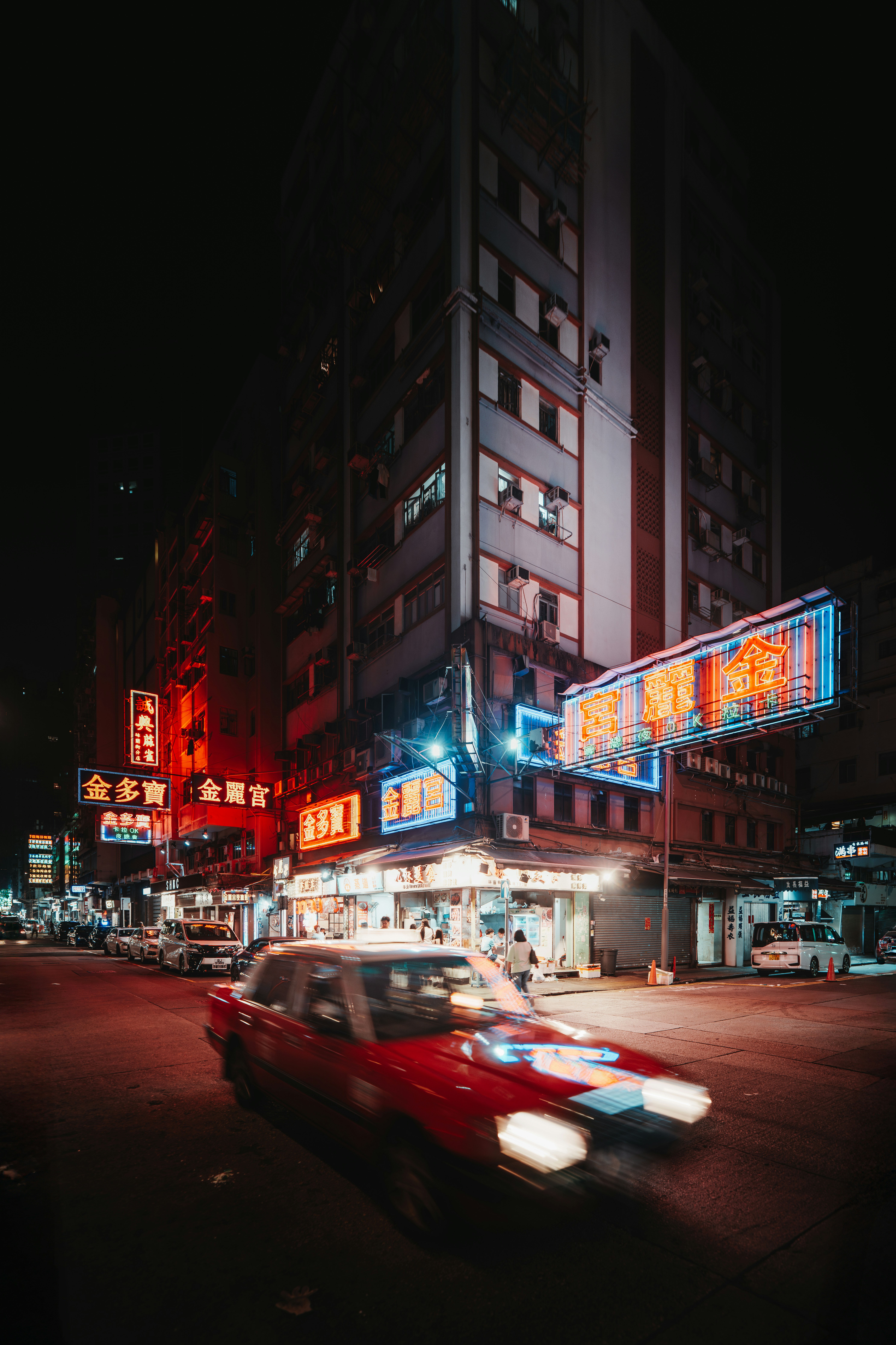 Vibrant neon signs illuminate a bustling street corner in a city at night, showcasing the lively atmosphere. A classic red taxi speeds by, adding to the dynamic scene.
