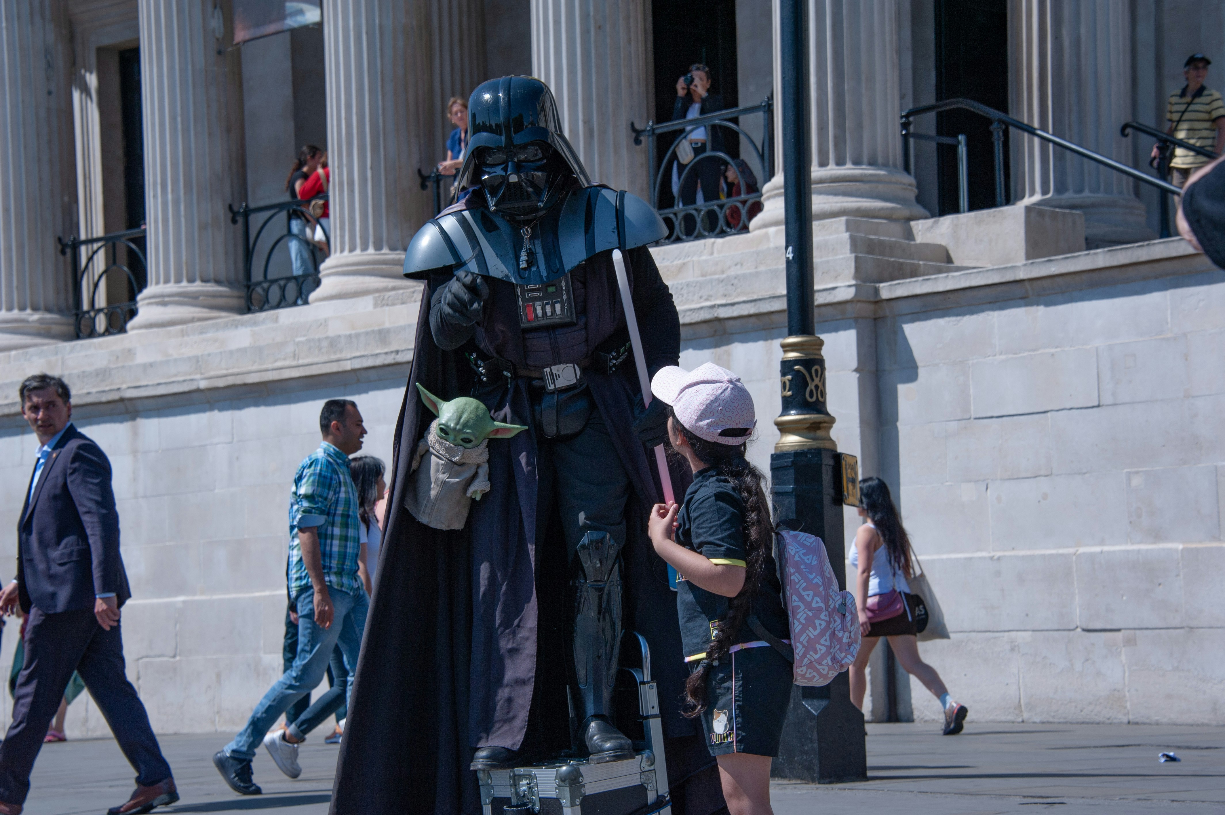 Darth Vader interacts with a young girl holding a toy Yoda in a bustling urban setting, surrounded by onlookers. The scene captures a playful moment blending pop culture with everyday life.