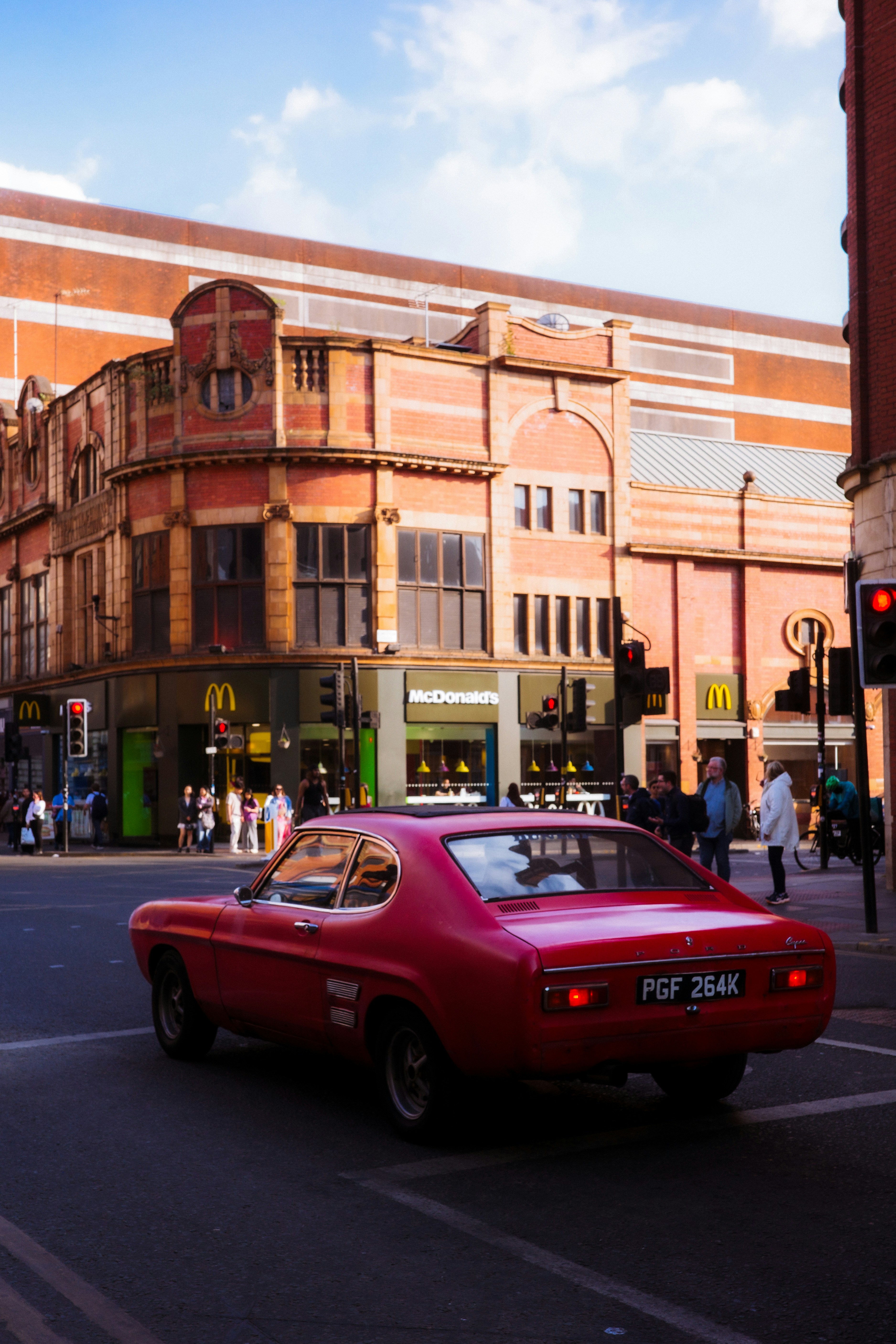 A vibrant pink classic car navigates a busy intersection near a McDonald's, showcasing a blend of vintage and contemporary urban life.
