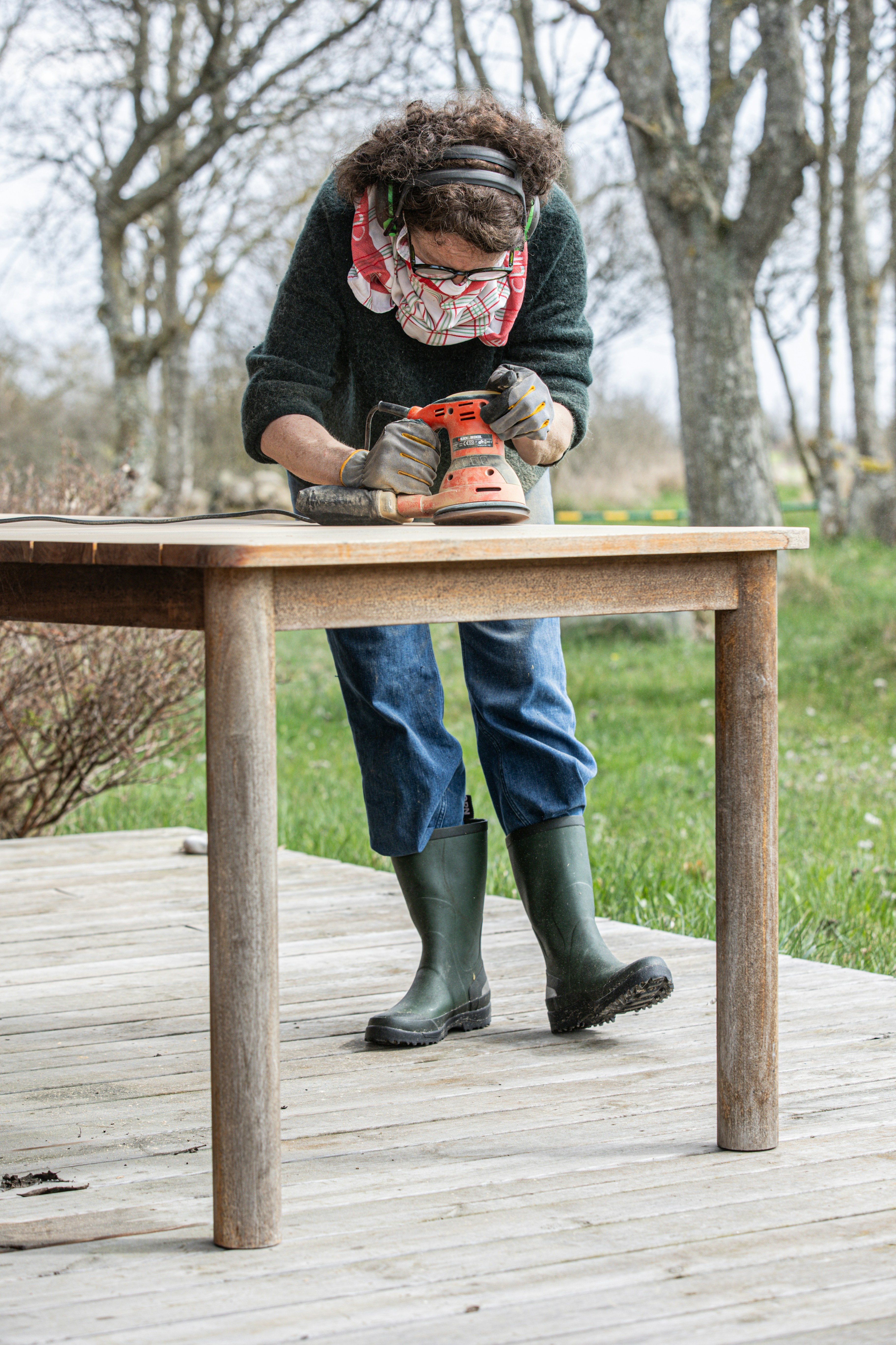 A person meticulously sanding a wooden surface on a sunny day, surrounded by trees and greenery. The scene captures the essence of craftsmanship and outdoor creativity.