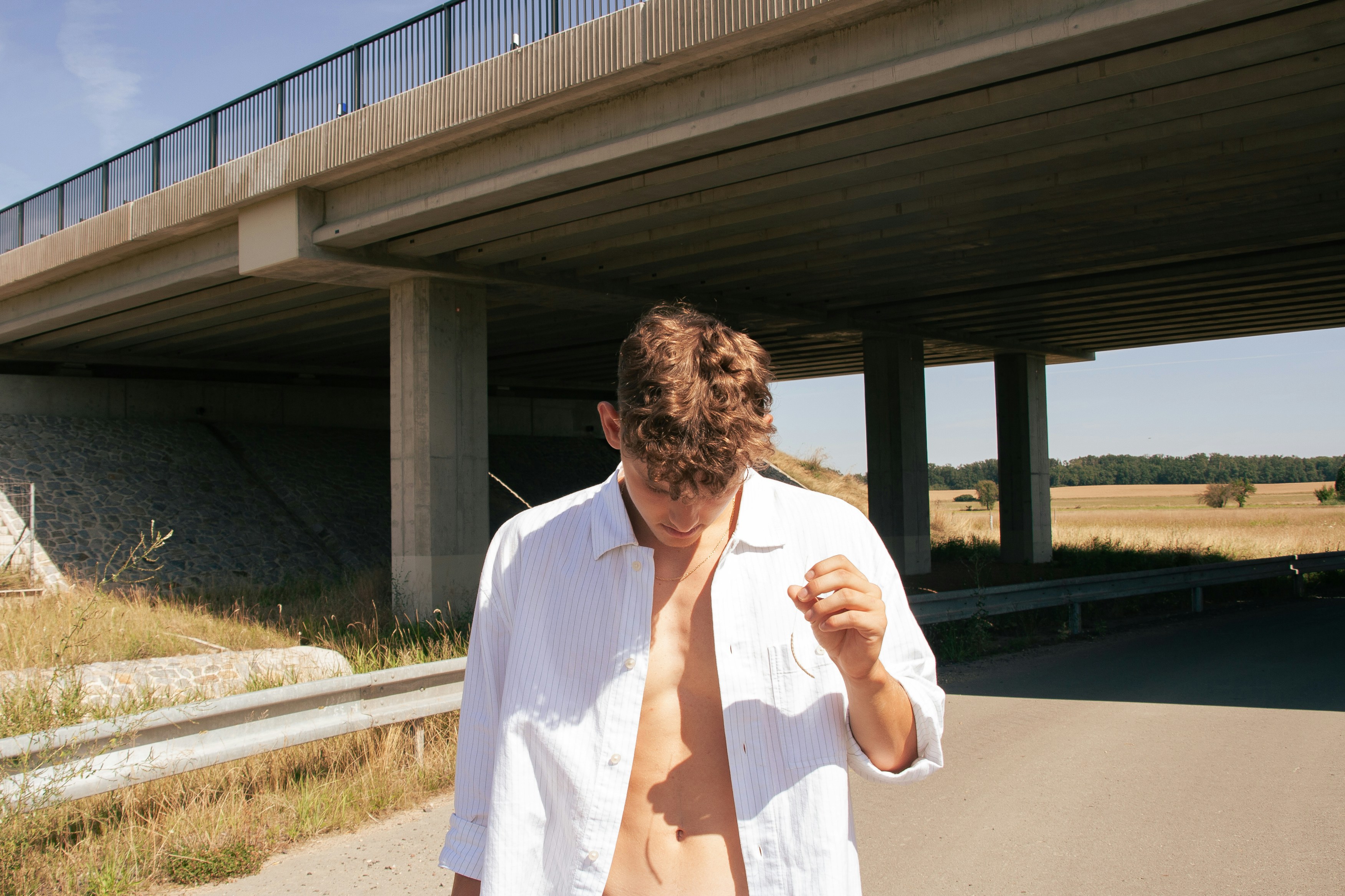 A man stands shirtless under an overpass, white shirt loosely draped, sunlit landscape in the background; the scene emphasizes casual vulnerability and urban openness.