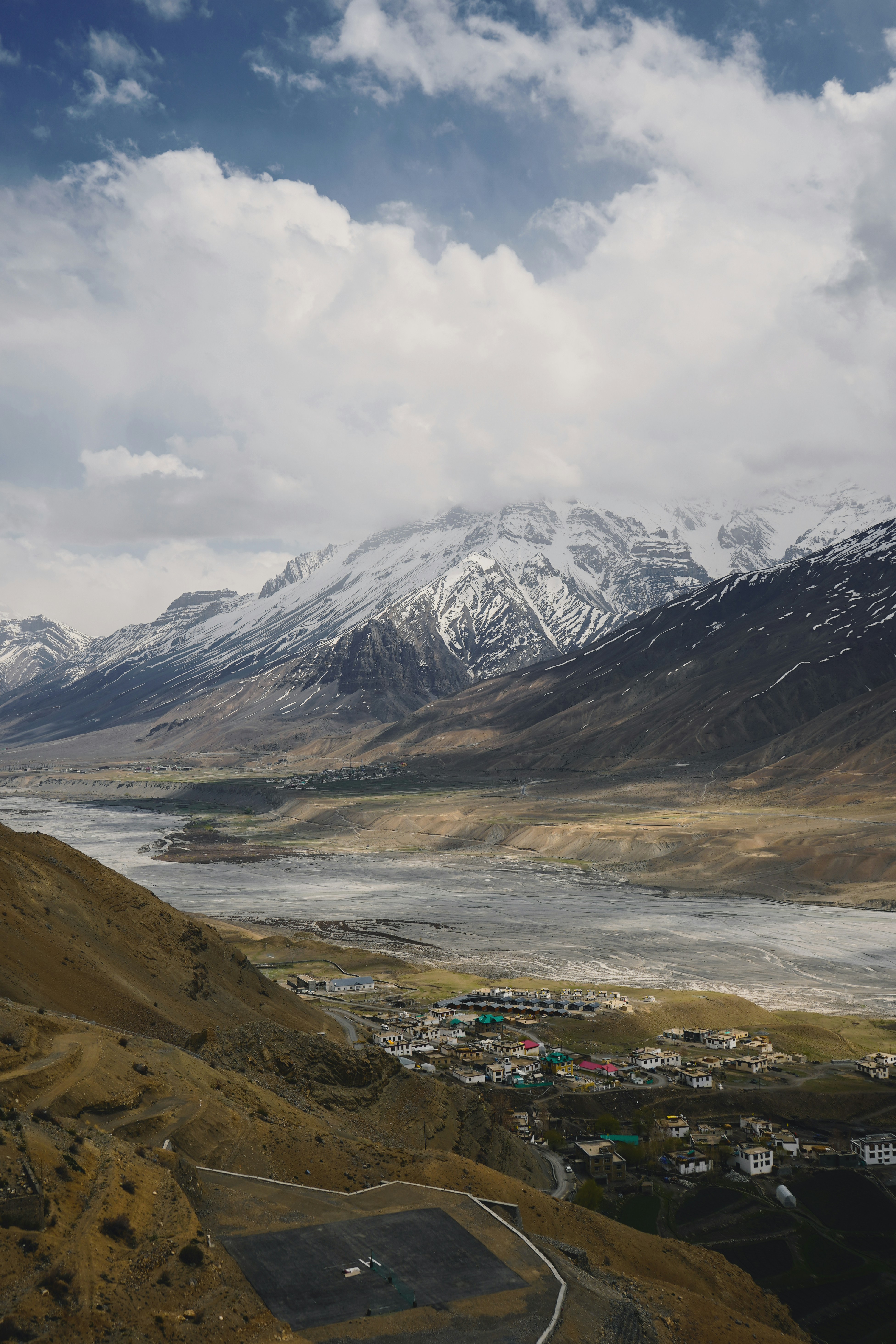 A panoramic view of a serene Himalayan valley, showcasing snow-capped peaks and a winding river alongside a small village. The dramatic sky adds depth to the landscape.
