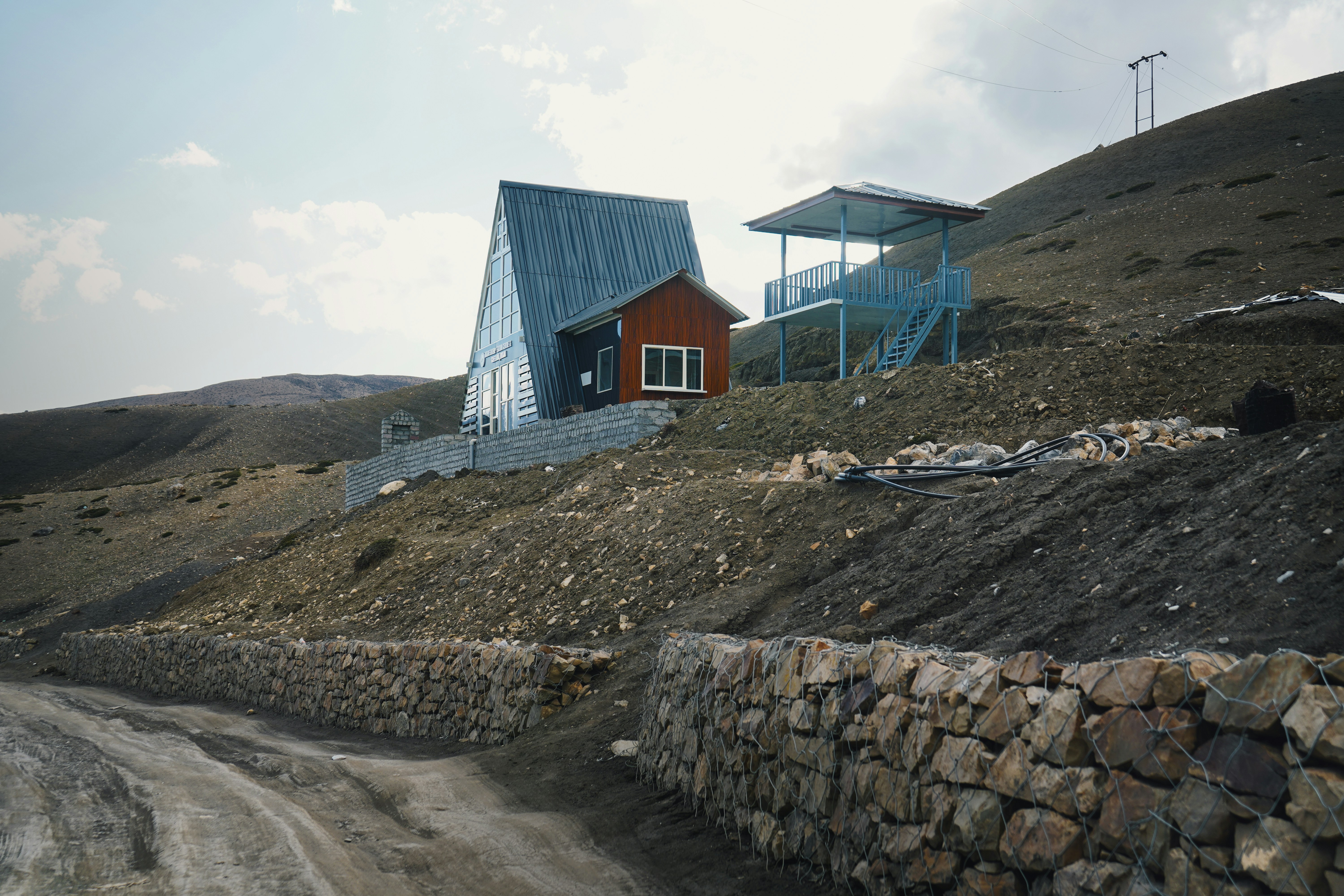 Modern house with a sloped roof and a deck, situated on a barren hillside with a rocky path leading up to it.