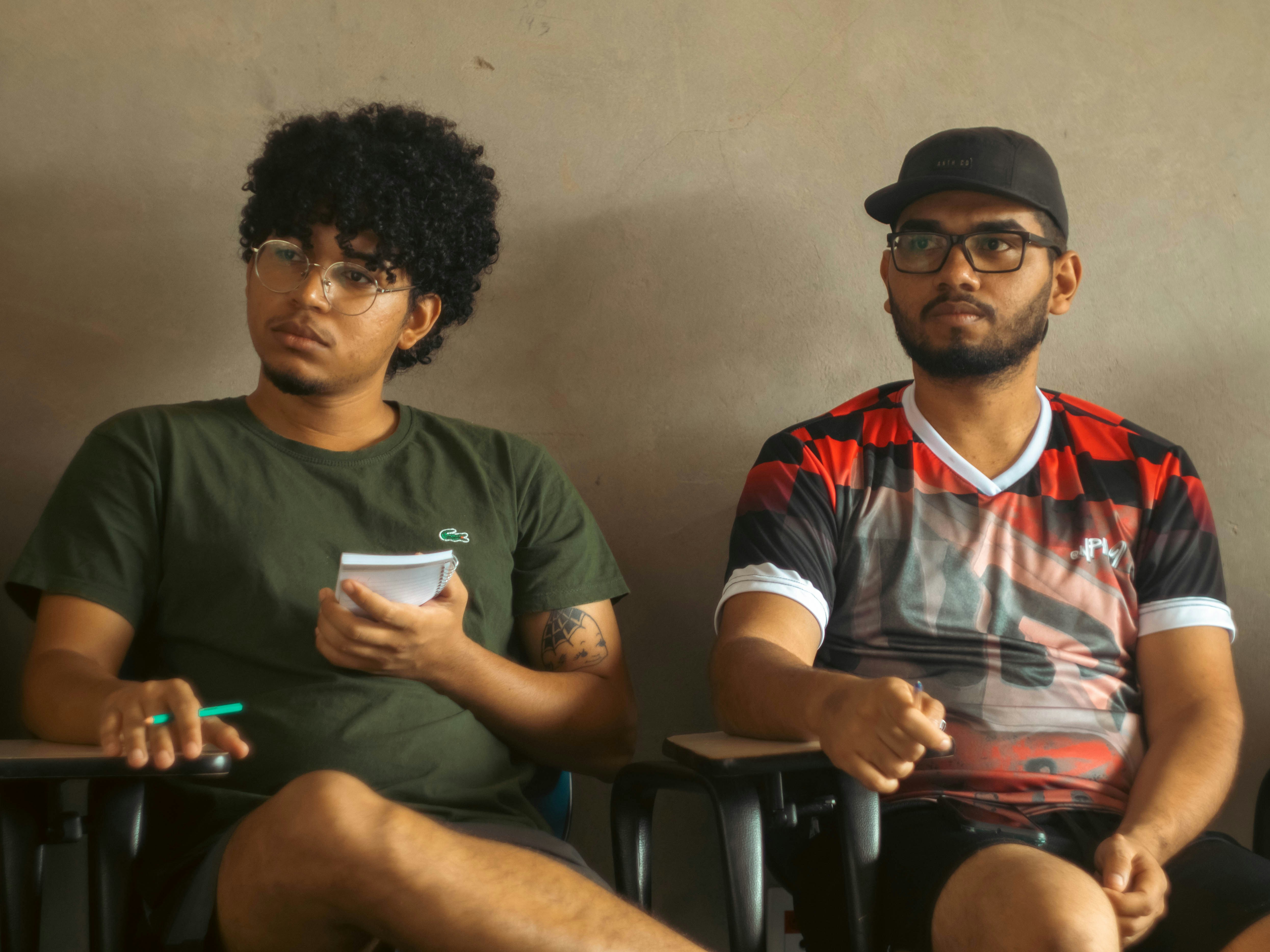 Two young men engaged in a thoughtful discussion during a workshop, with one holding a notepad. The setting suggests a collaborative learning environment.