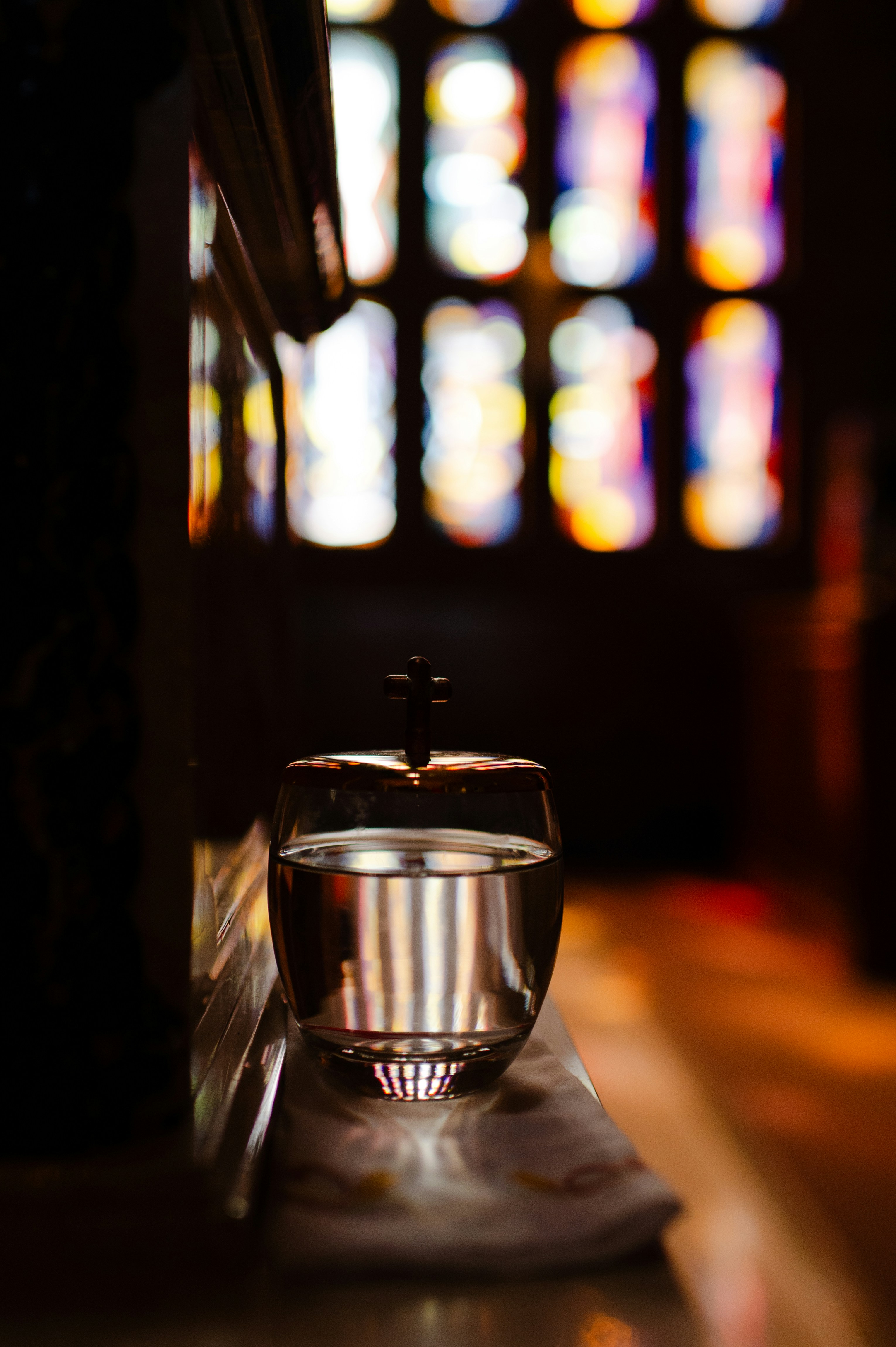 A single glass of water rests on a ledge inside a dim, candlelit church, with blurred stained glass creating warm bokeh in the background.