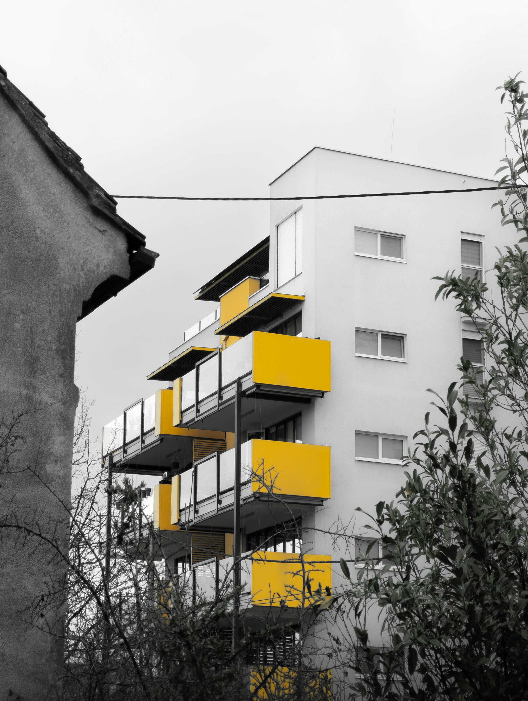 Modern apartment building with striking yellow balconies contrasted against a black-and-white background of an older structure and foliage.
