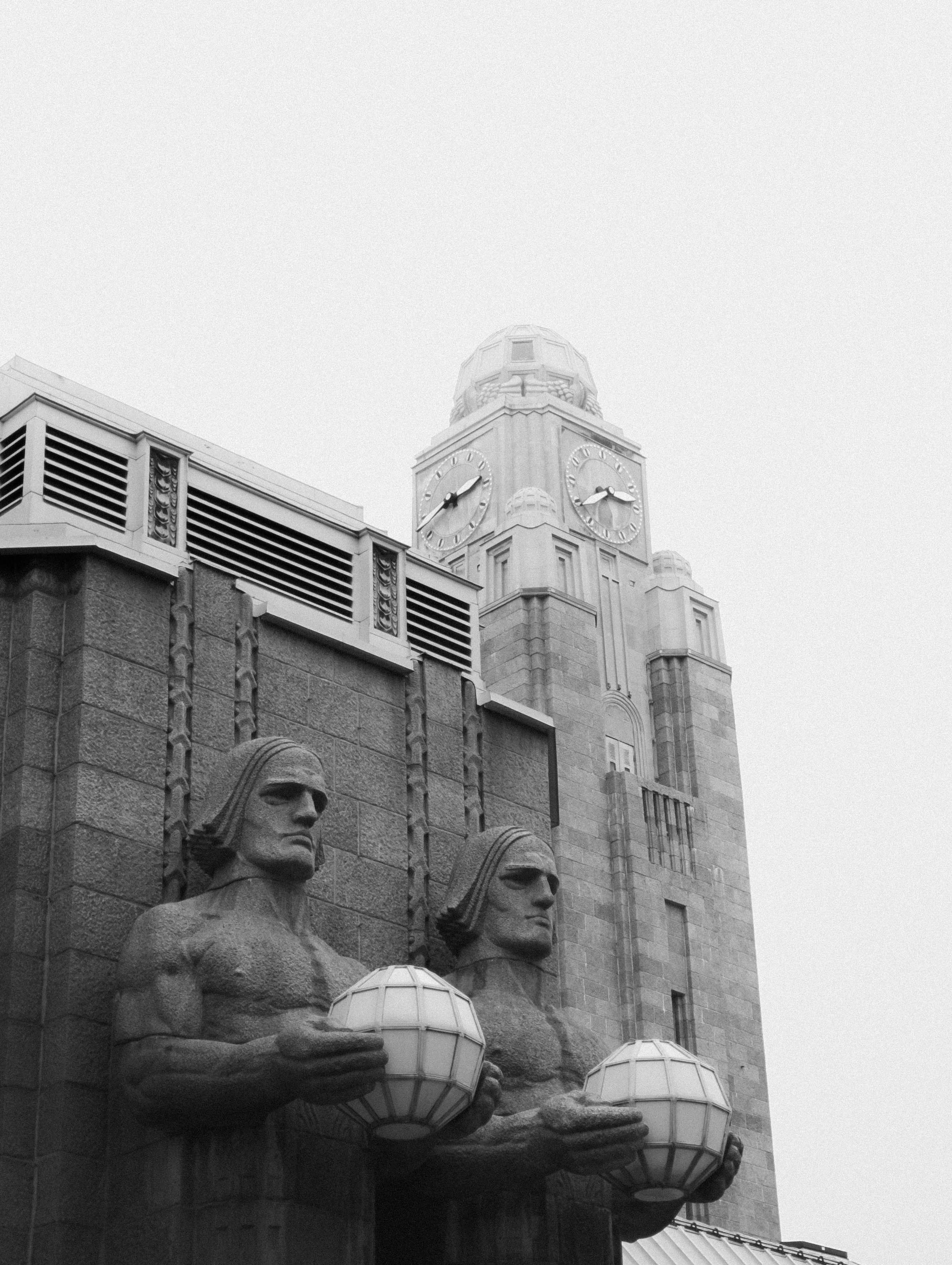 Two imposing statues hold luminous orbs in front of a historic clock tower, enveloped in a misty atmosphere.