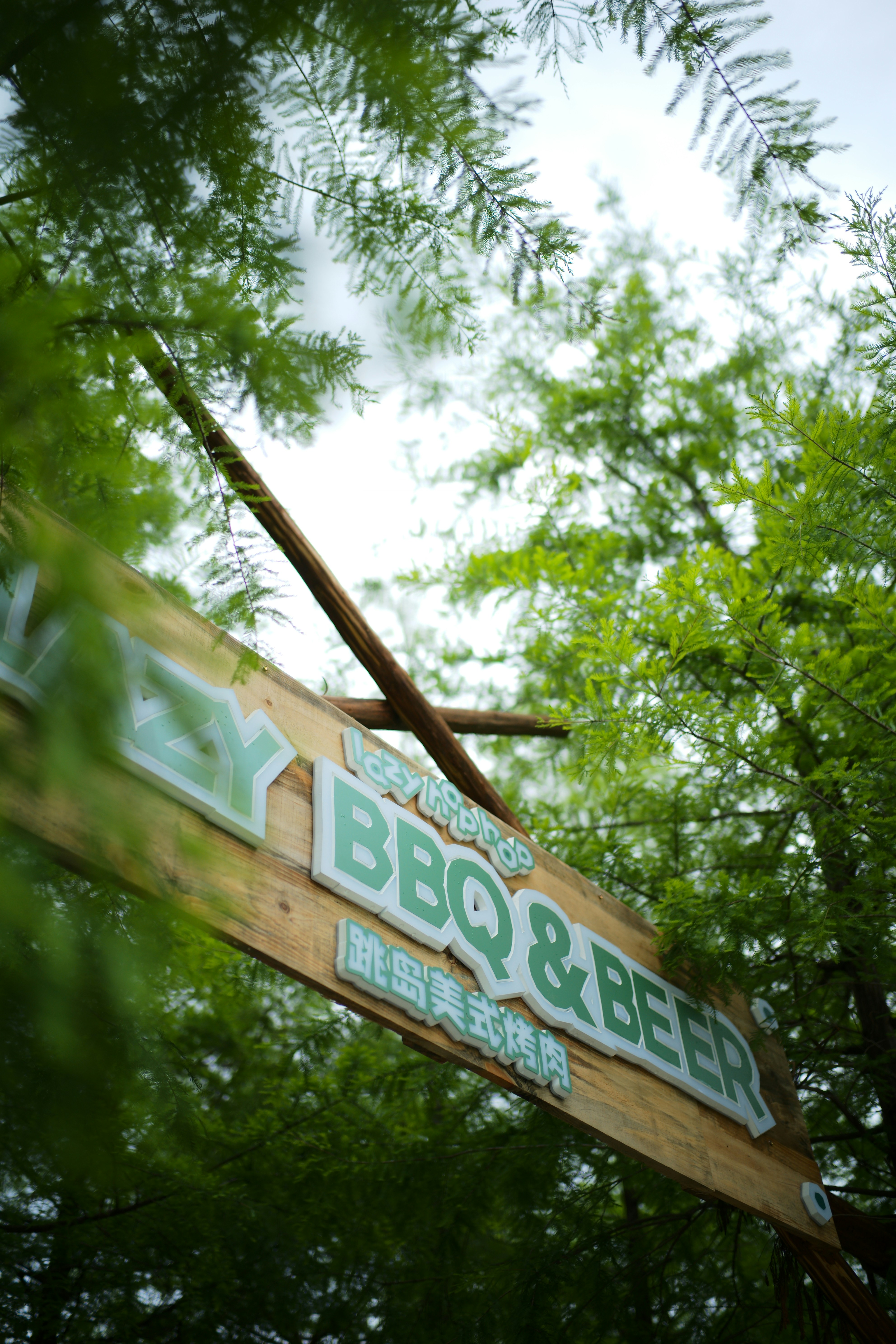 Wooden sign for a BBQ and beer venue, partially obscured by lush greenery. The sign features playful typography and vibrant colors.