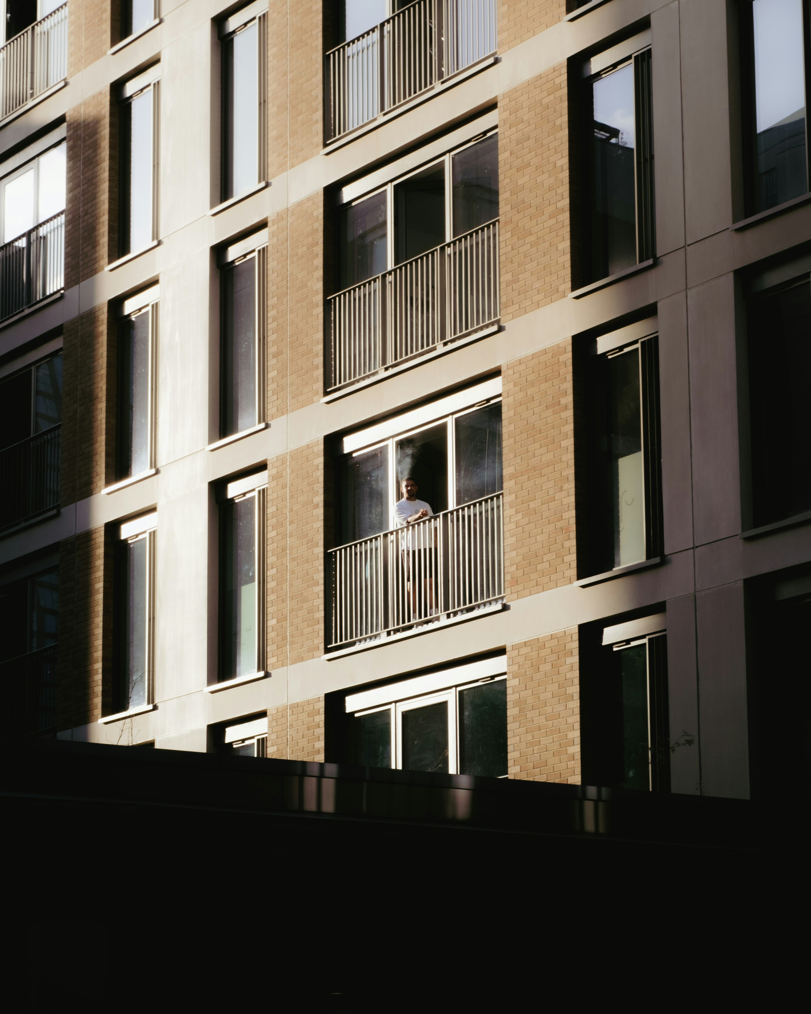 Person standing on a balcony of a modern apartment building, framed by sunlight and shadows. The architecture contrasts with the figure's stillness.