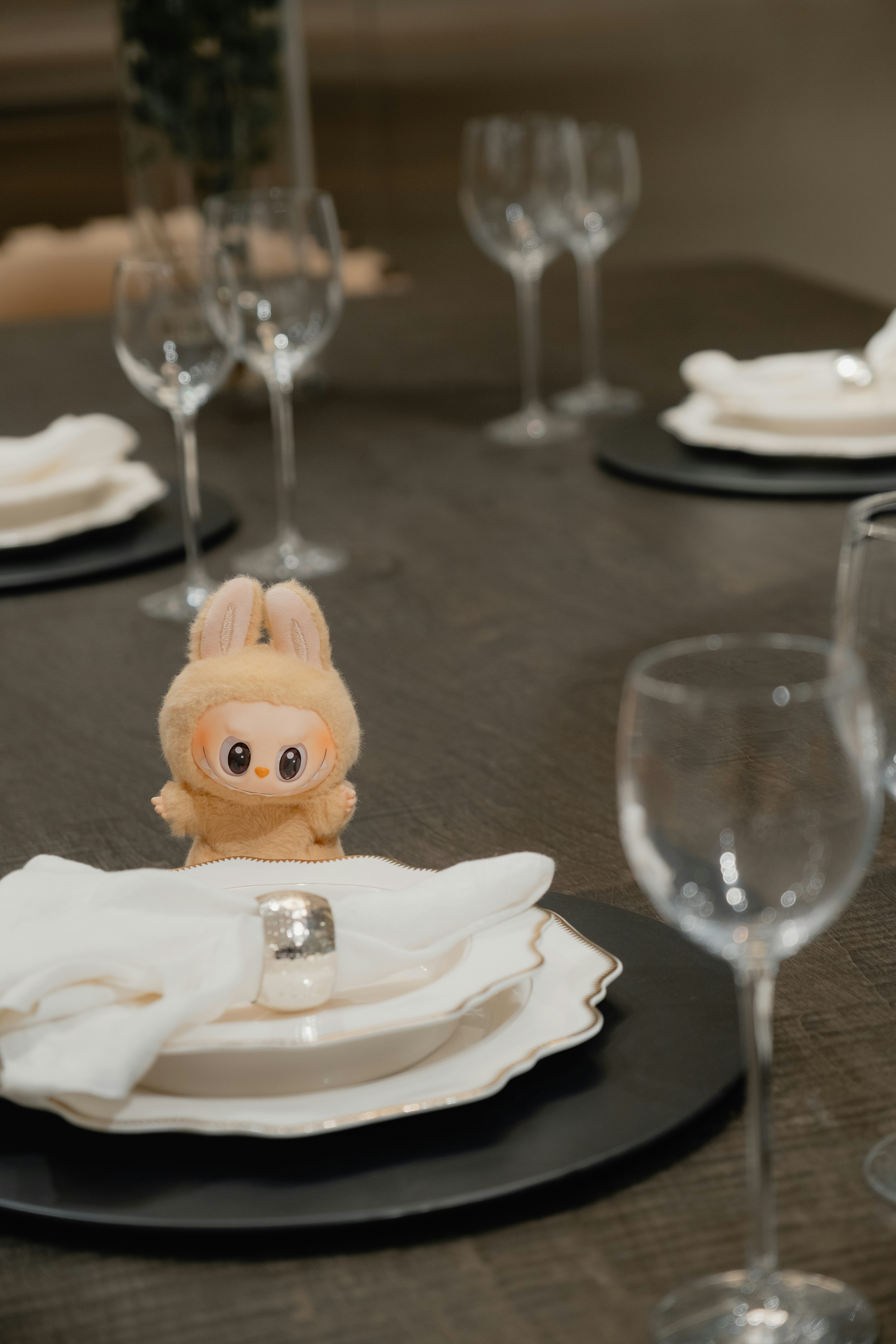 A small plush bunny sits on a white napkin atop a plate at a formal dining table, surrounded by wine glasses.