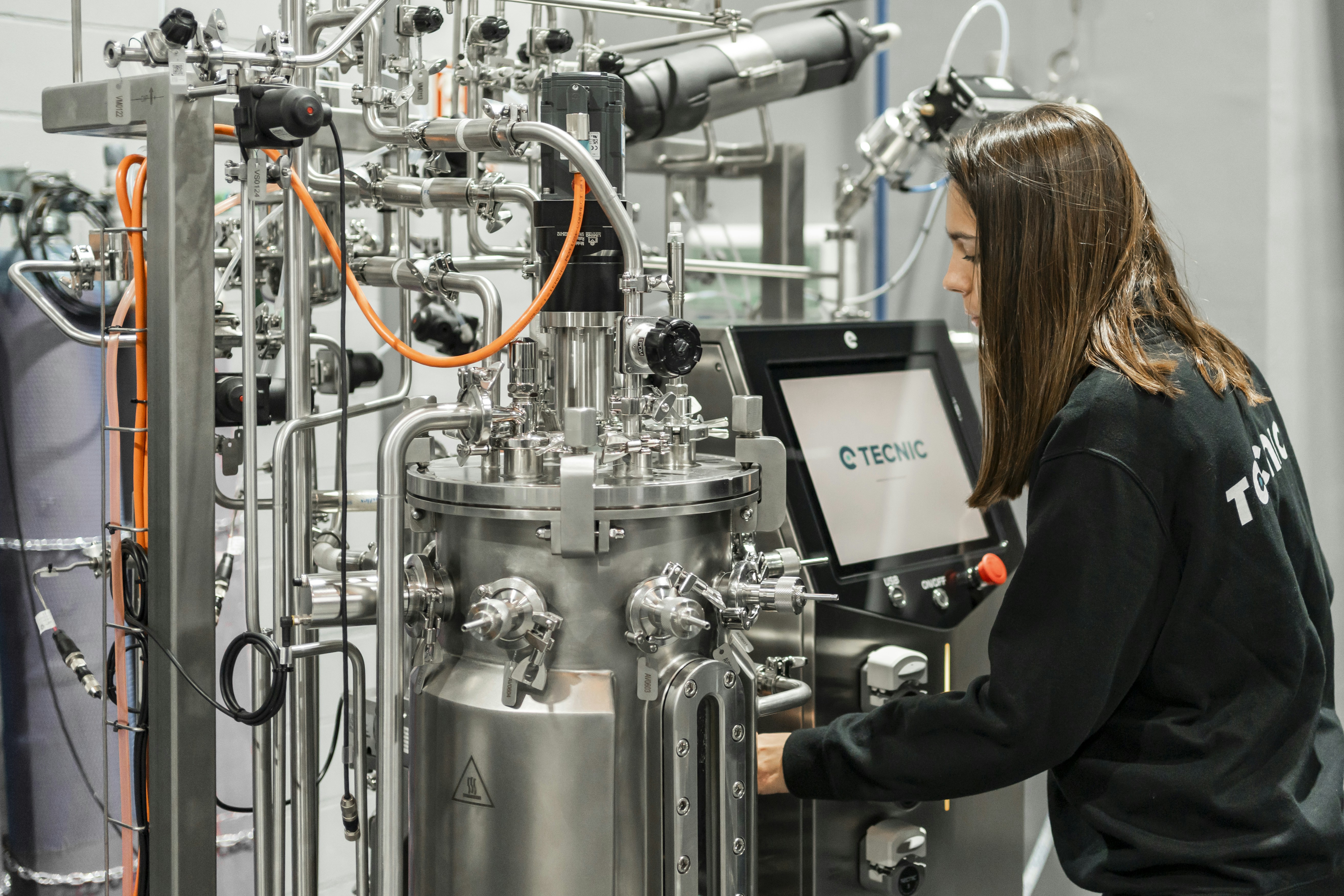 Operator handling a stainless steel bioreactor with integrated touch screen in a technical process control environment.