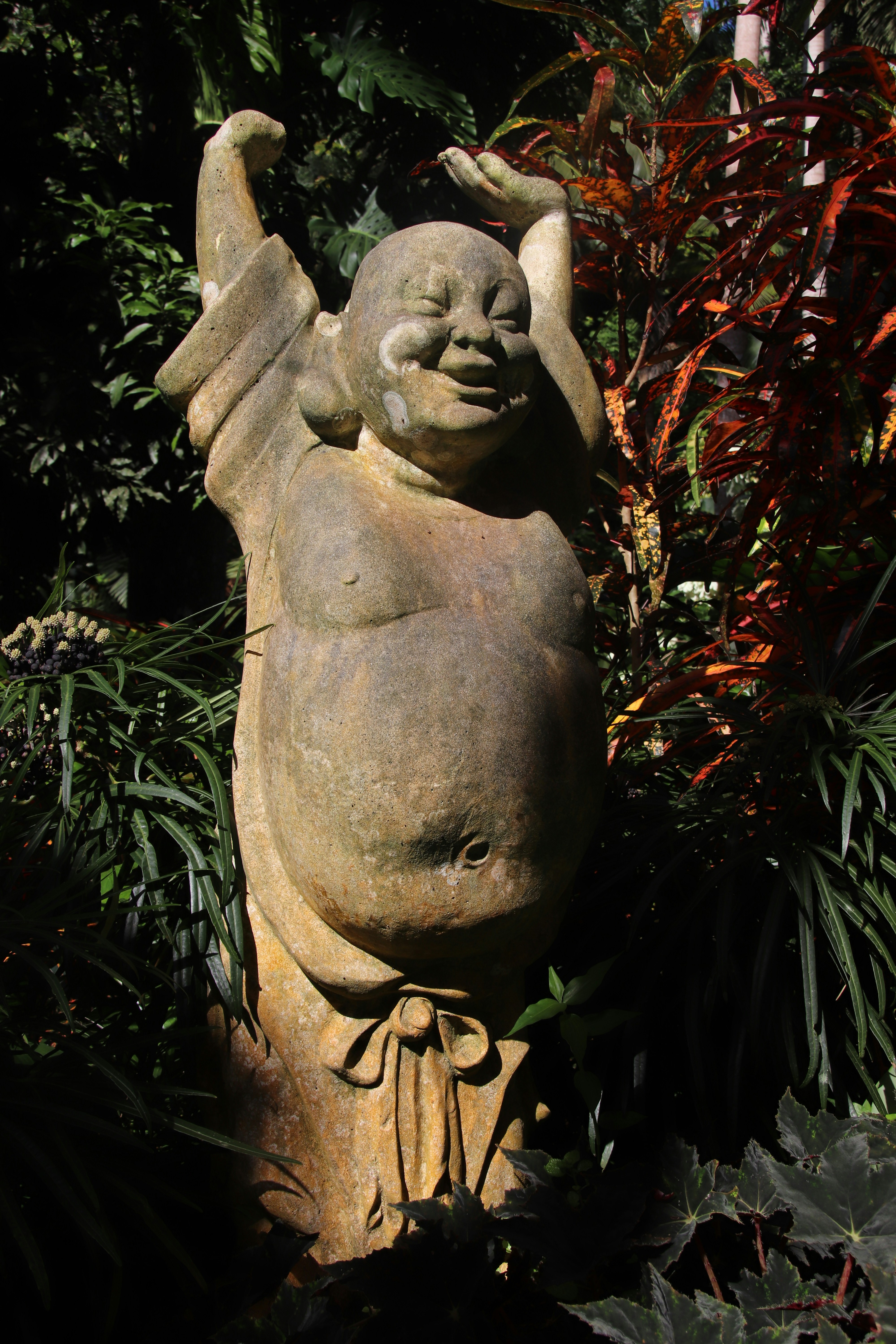 A cheerful stone statue with raised arms stands amidst vibrant foliage, exuding a sense of happiness and tranquility.