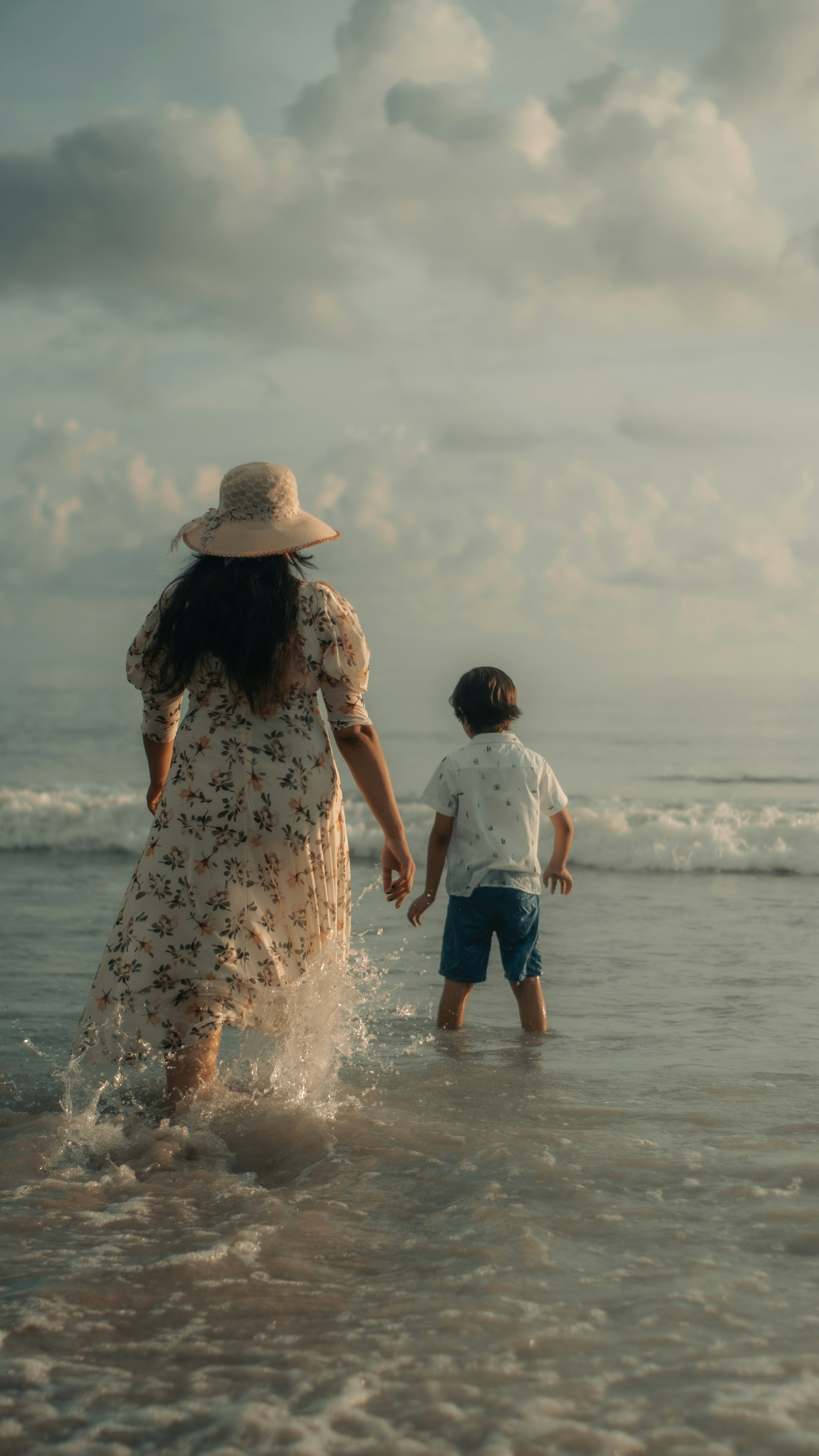 A woman in a floral dress and a hat walks hand-in-hand with a child through gentle ocean waves, capturing a moment of joy and connection at the beach.