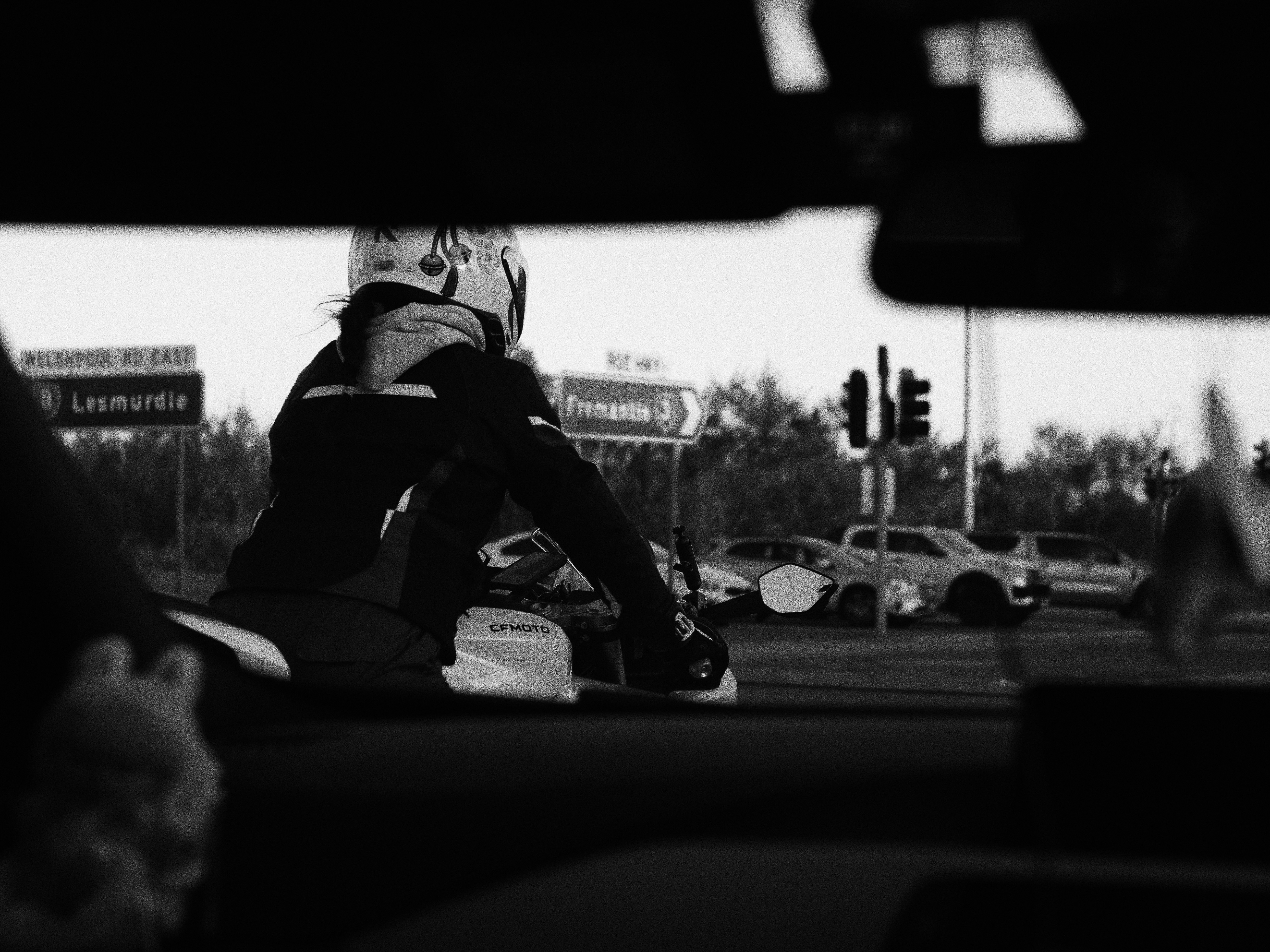 Motorcycle rider waiting at a traffic signal, framed within a vehicle's interior. Road signs and other vehicles create a dynamic urban backdrop.