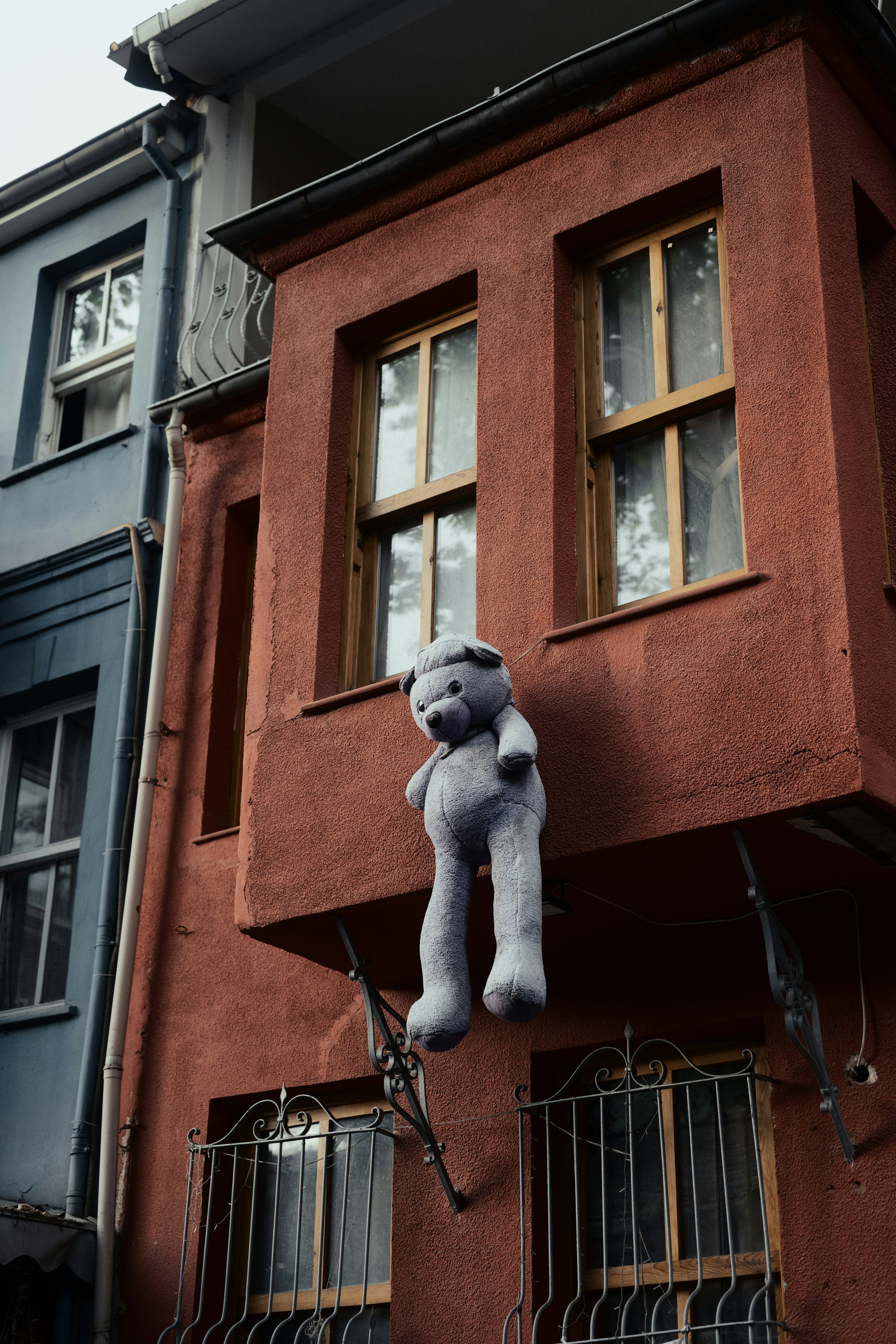 A plush teddy bear hangs from a balcony of a colorful building, evoking a sense of nostalgia and whimsy. The contrasting colors of the building add to the visual intrigue.