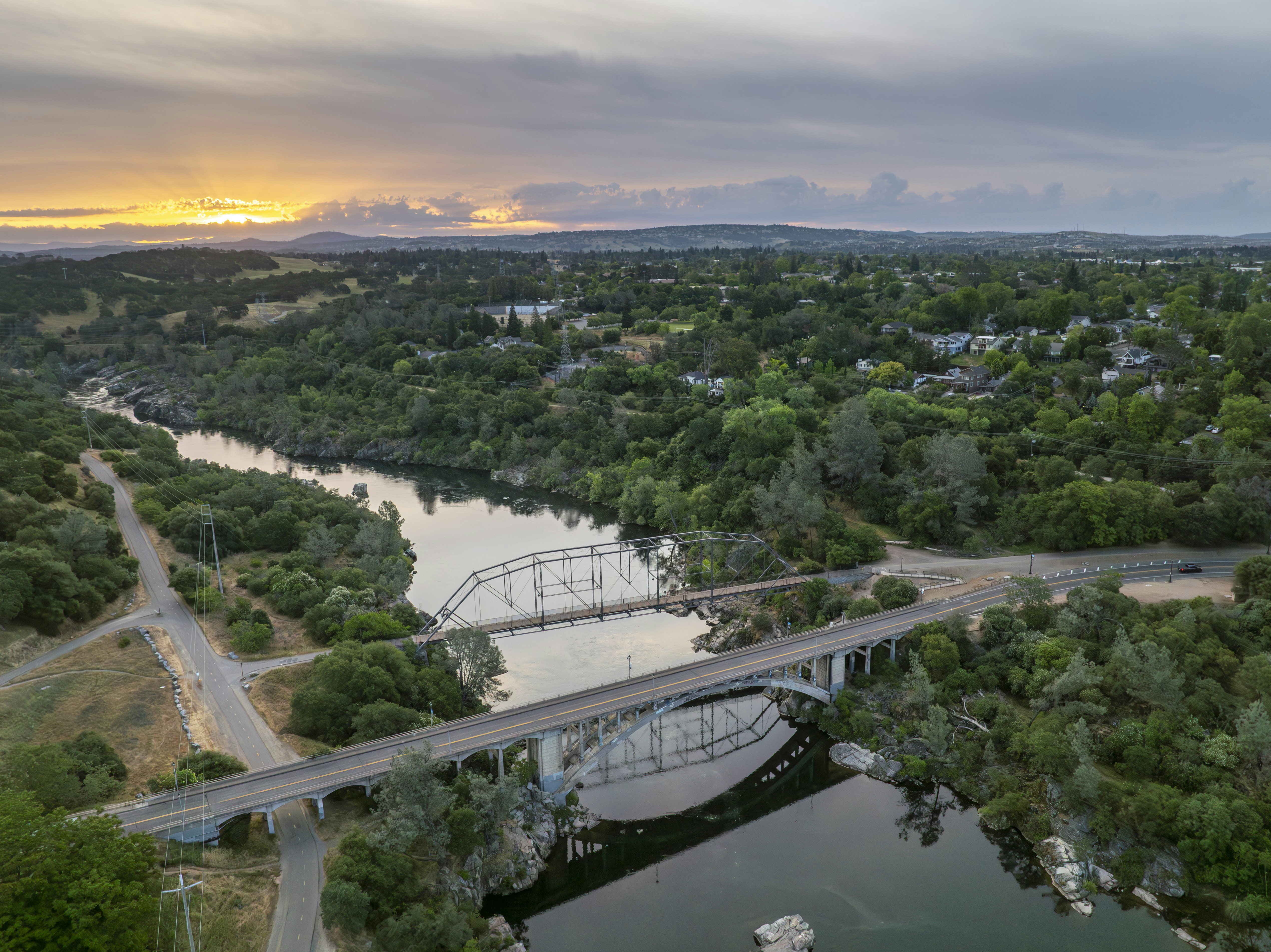 Aerial view capturing two bridges spanning a serene river, framed by lush greenery and a colorful sunset sky.