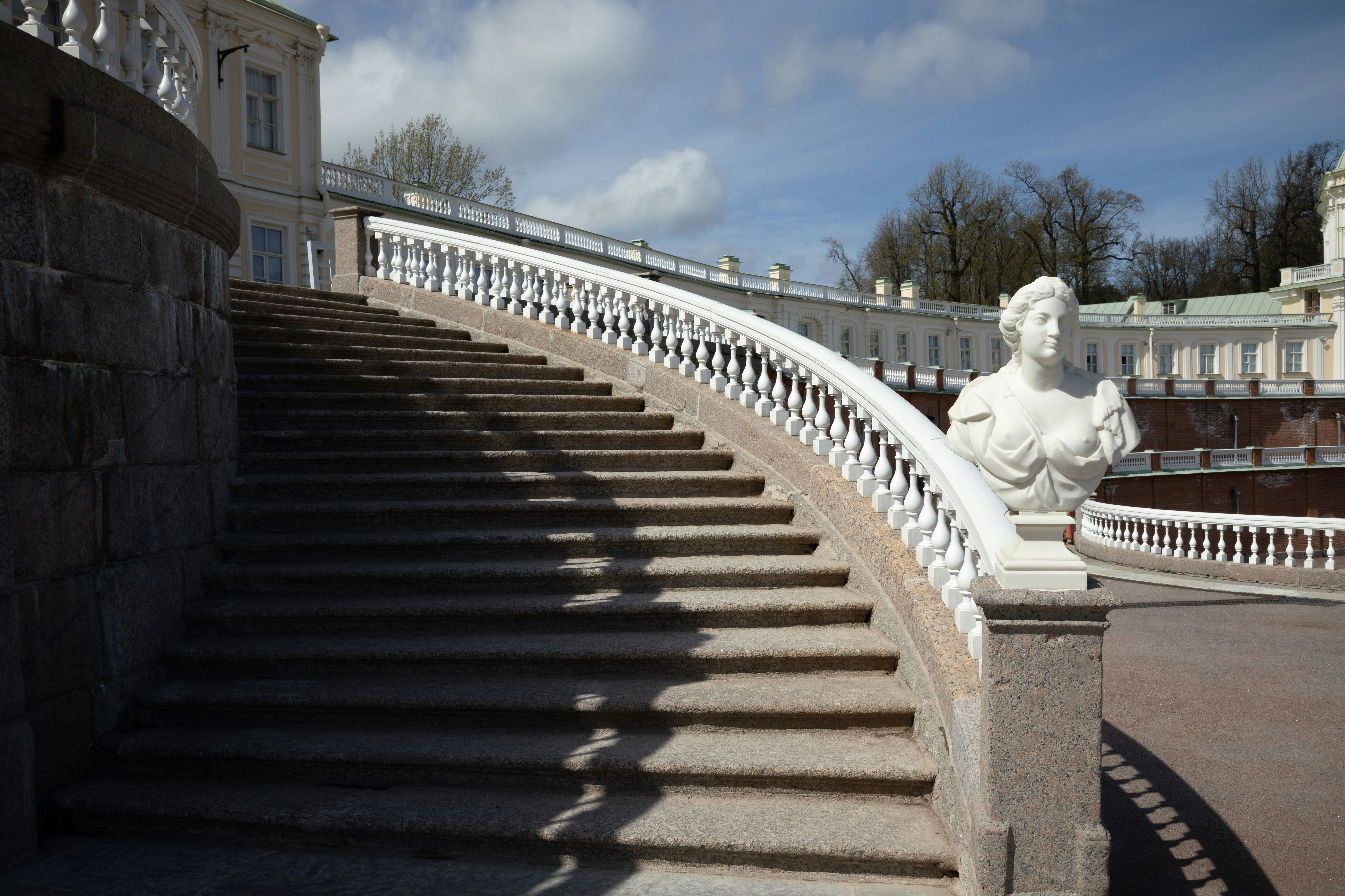 Graceful staircase leading to a classical bust, framed by a grand architectural backdrop and soft clouds above.