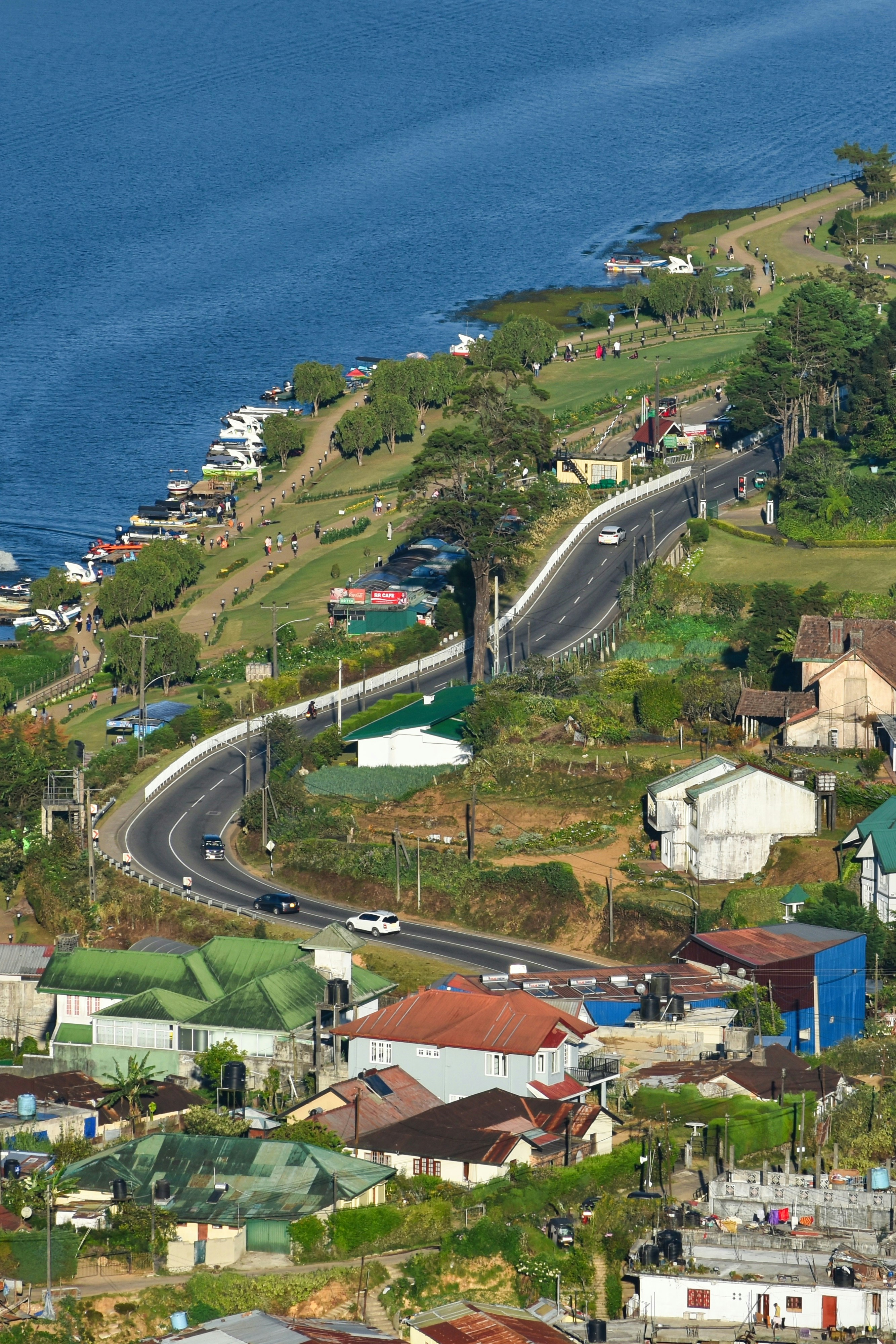 Aerial view of a winding road alongside a serene lake, surrounded by lush greenery and scattered buildings.