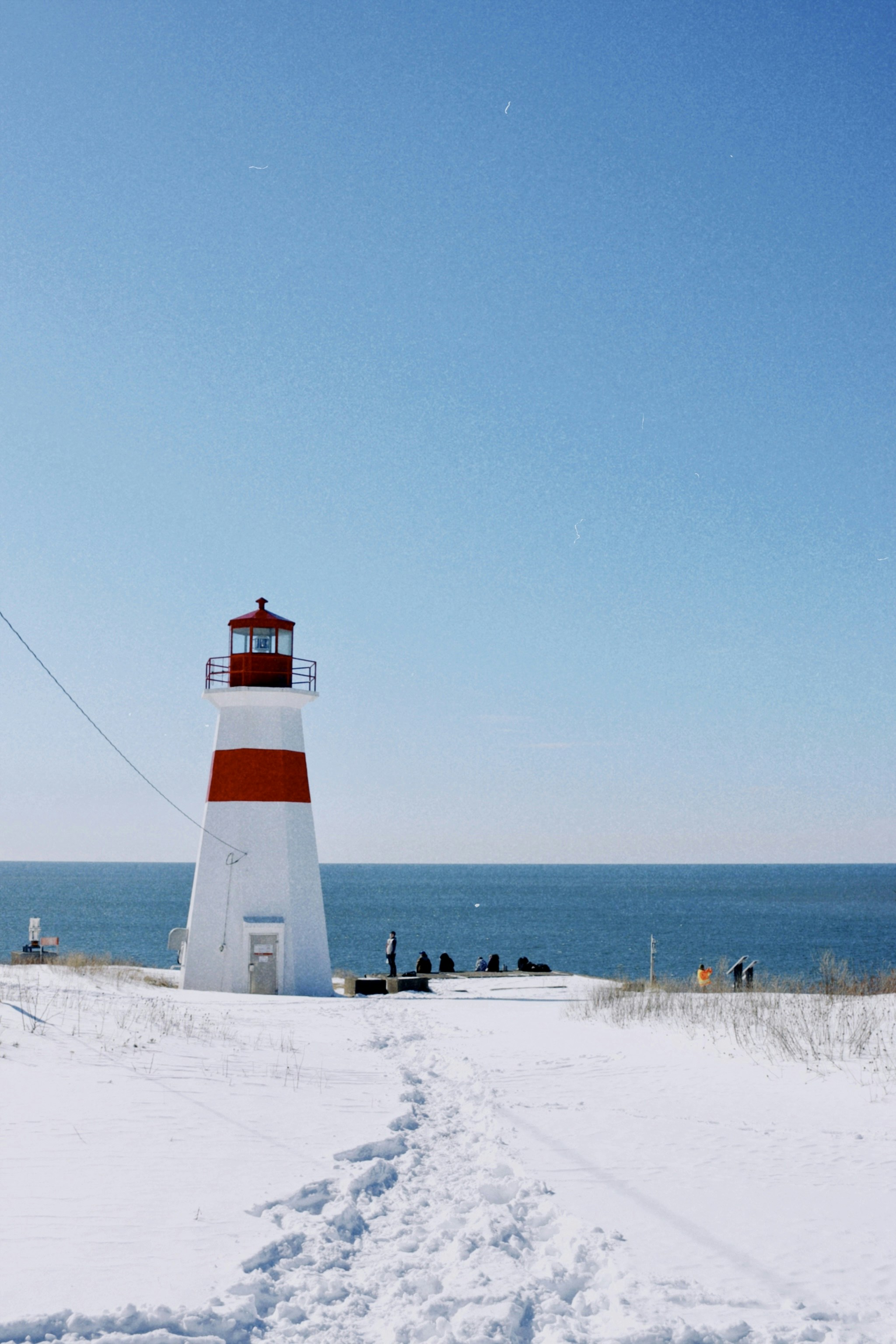 A red and white lighthouse stands tall against a serene ocean backdrop, surrounded by snowy terrain and a clear blue sky.