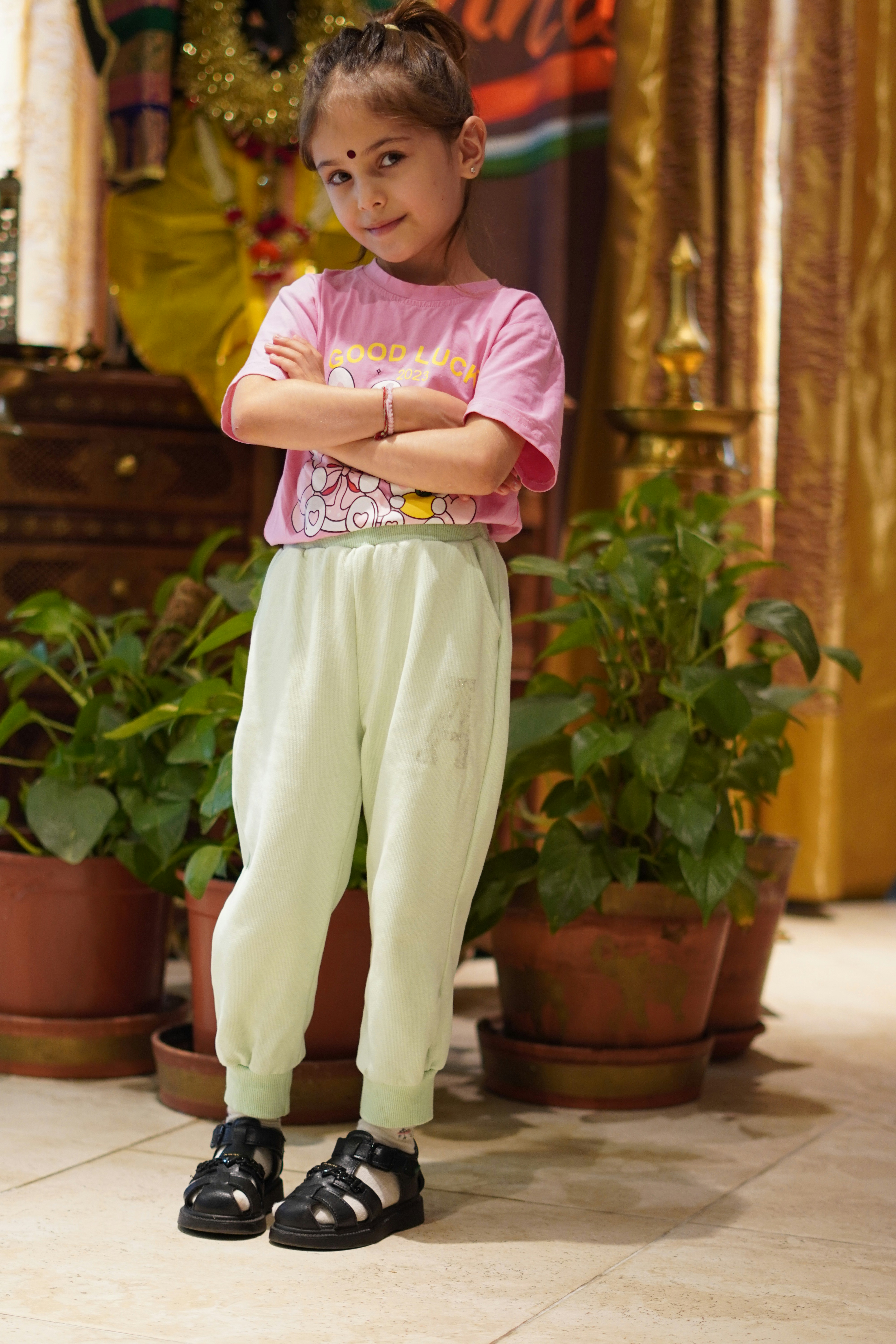 Young girl with a playful expression stands confidently among potted plants, dressed in a pink graphic tee and light green pants. The warm backdrop enhances the vibrant scene.