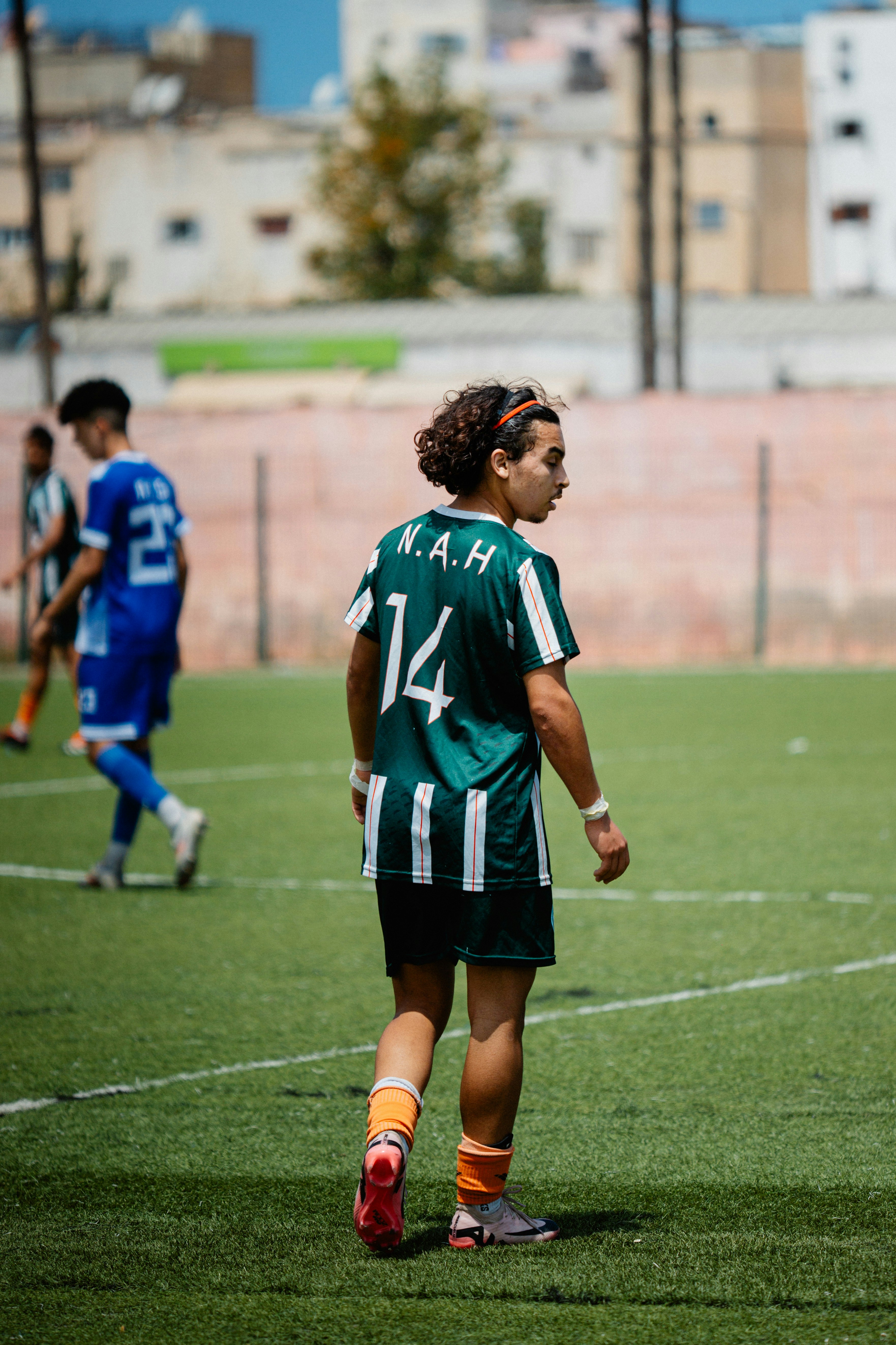 A young soccer player in a green and white jersey with 'N.A.H.' and the number 14 walks across a vibrant green pitch, showcasing focus amidst teammates in the background.