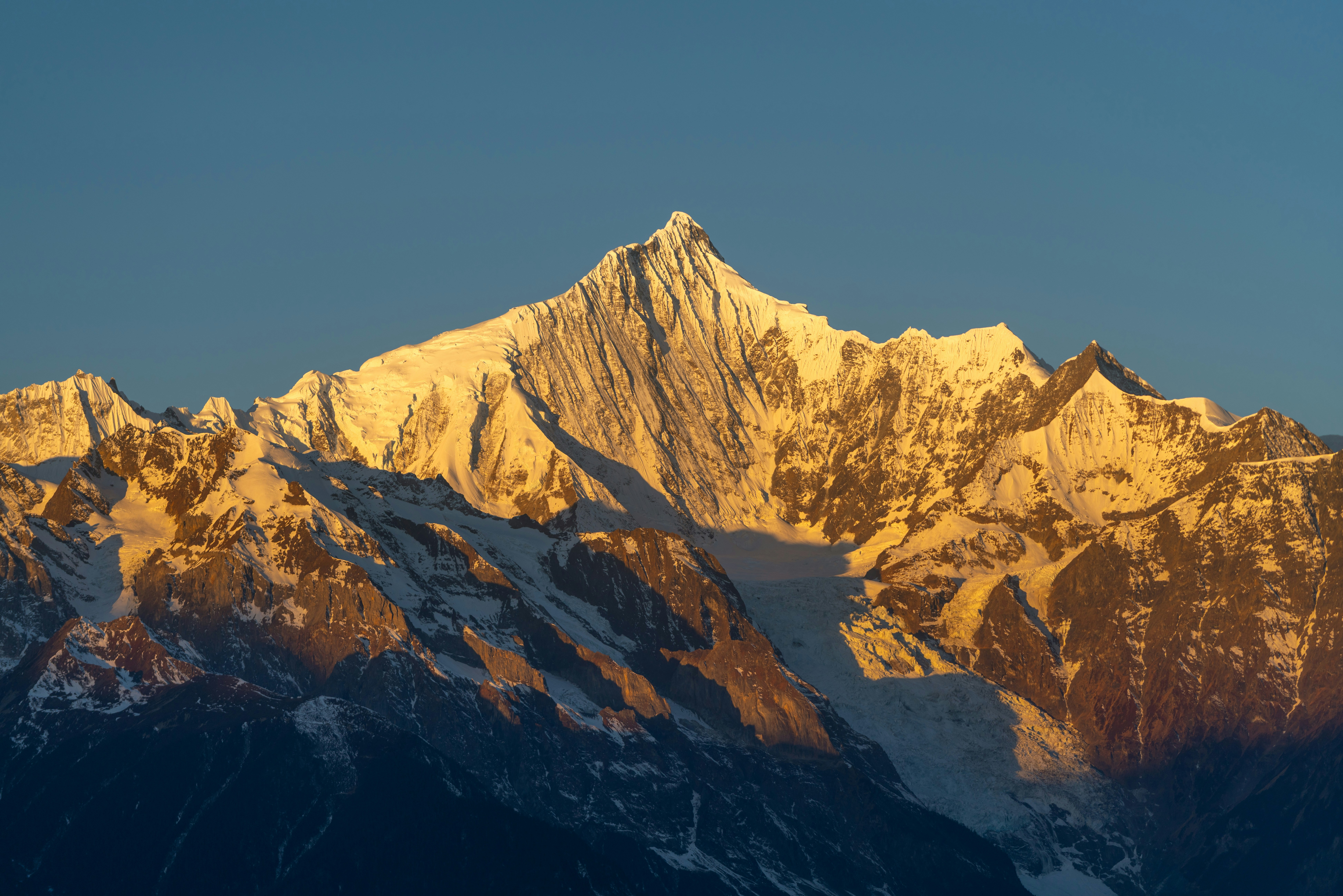 Snow-capped mountain peak illuminated by golden sunlight against a clear sky. The rugged terrain showcases the majesty of nature's architecture.