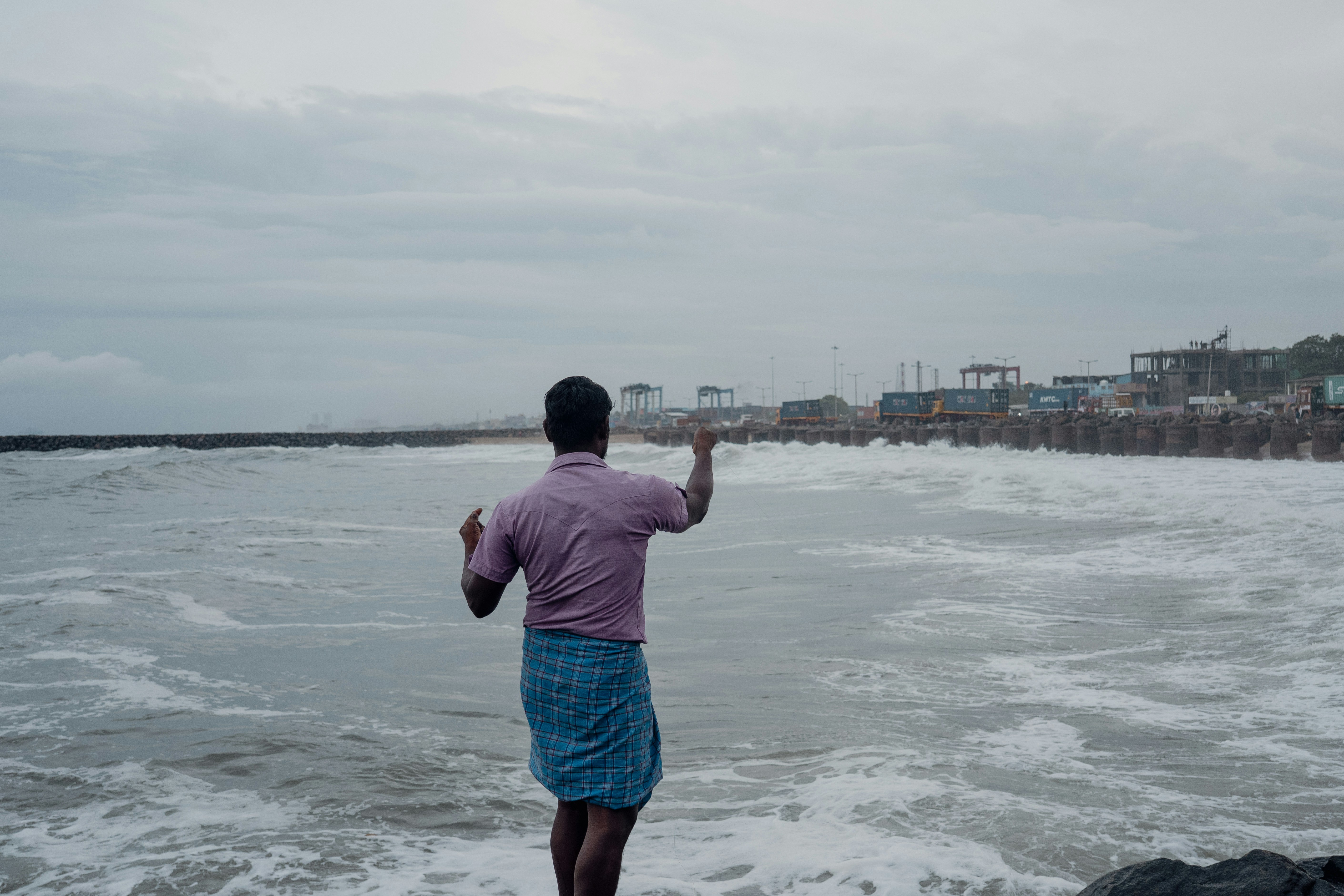 Man standing by the shore, gesturing towards the ocean as waves crash around him. The scene captures a moment of contemplation against a backdrop of a cloudy sky.