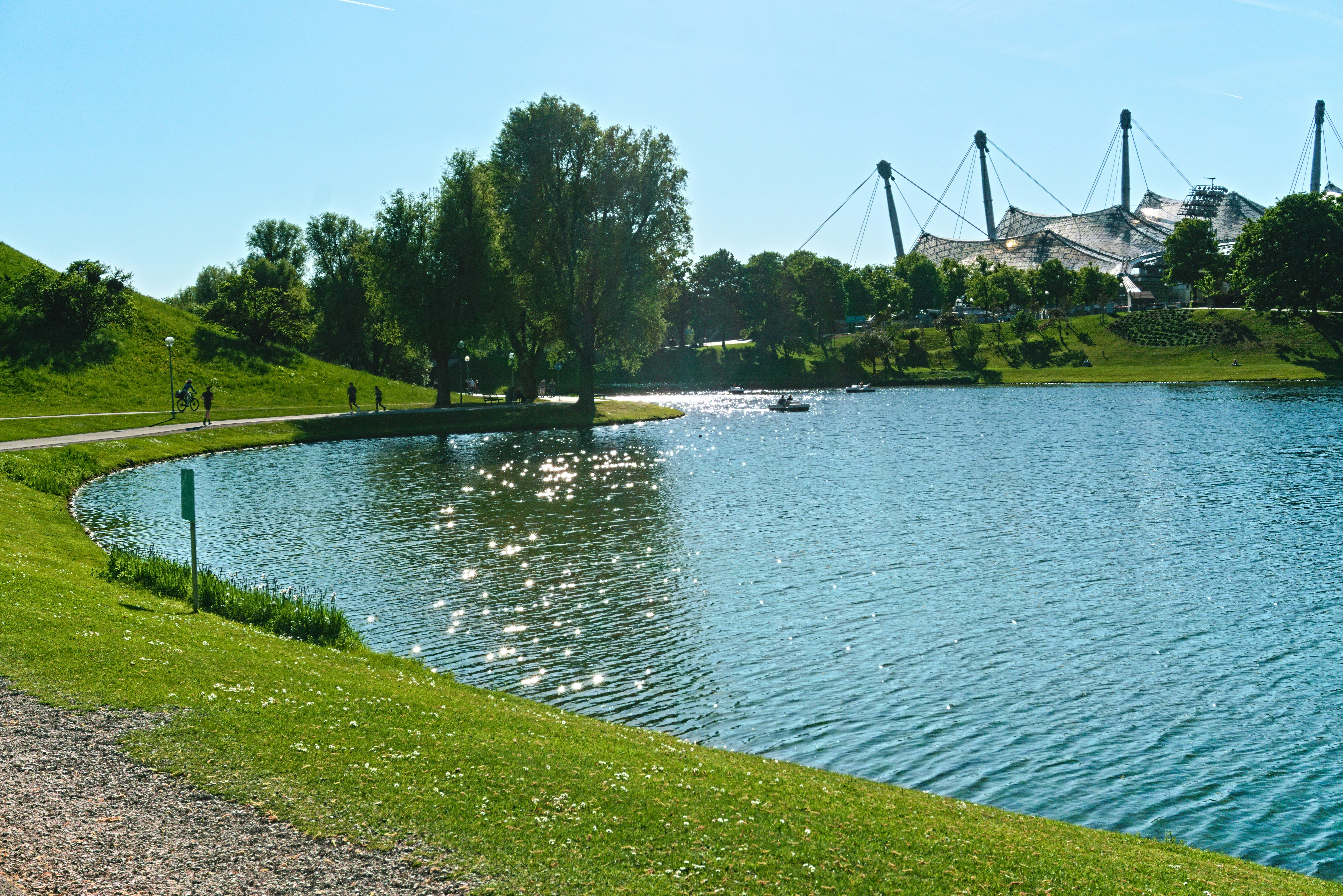 Lush green park with a winding path alongside a shimmering lake, featuring distant structures and recreational activities. 