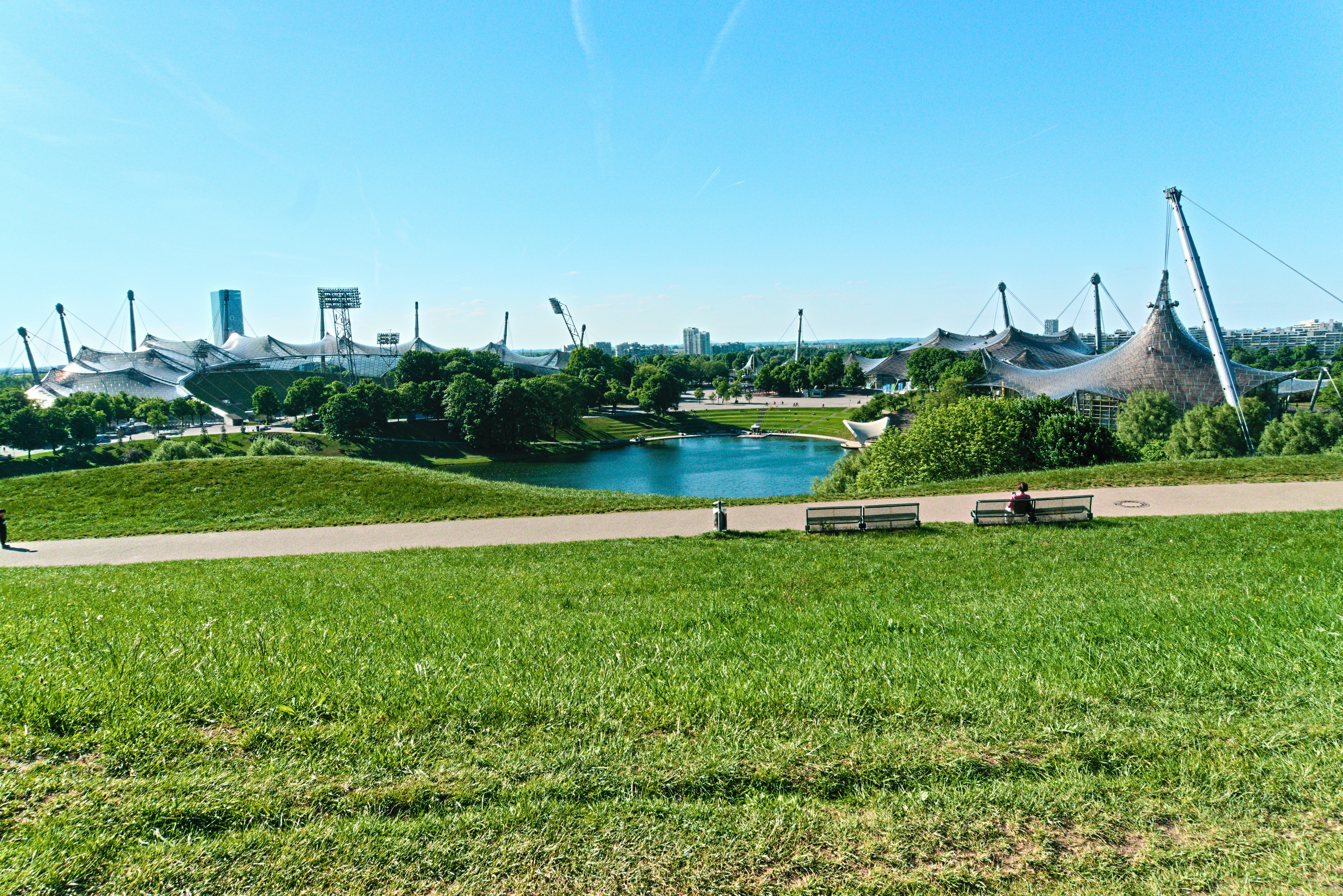 Lush green park with a serene pond surrounded by iconic structures and gentle hills. A couple sits on a bench, enjoying the tranquil atmosphere.